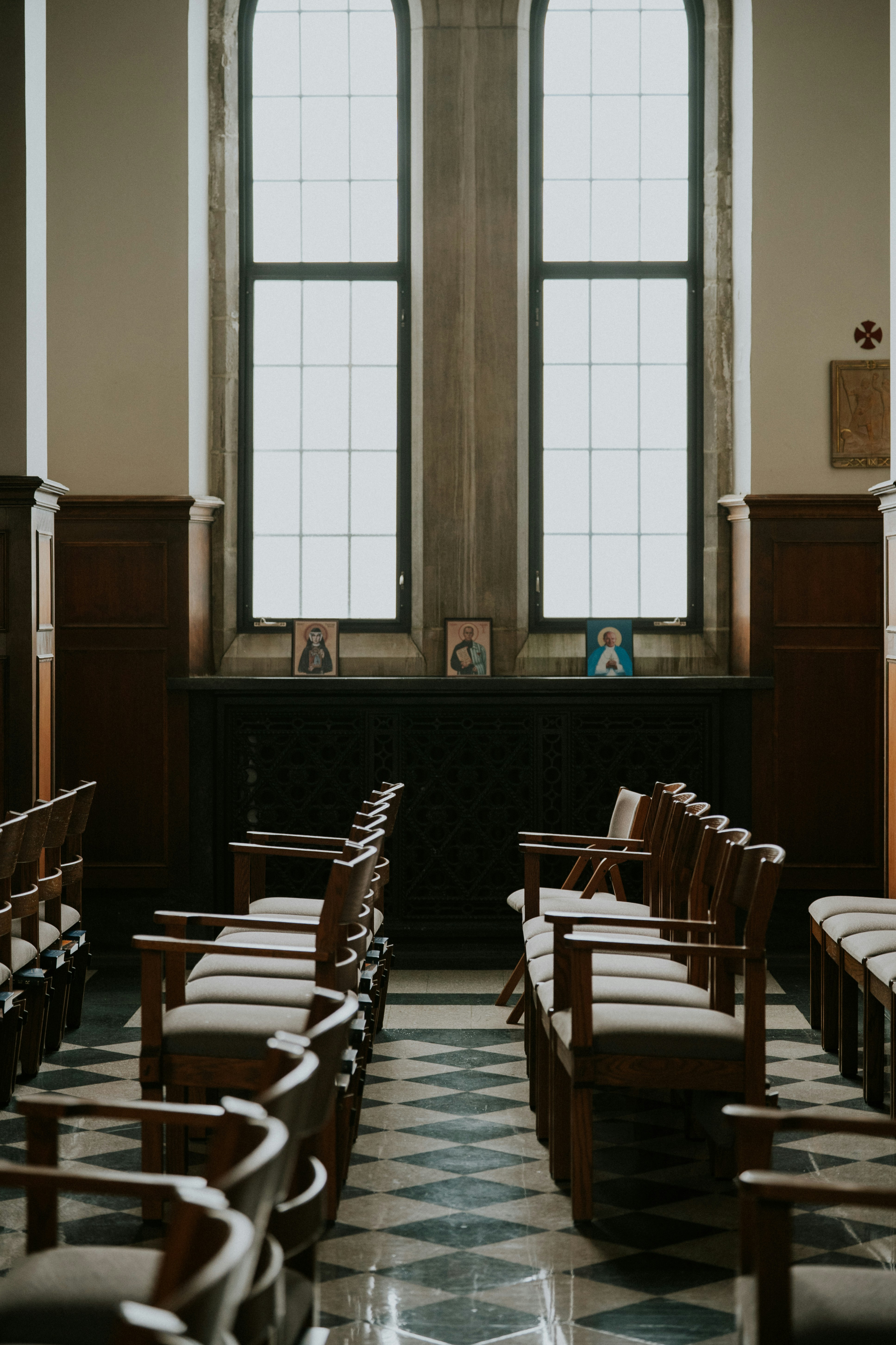 A quiet chapel interior featuring rows of wooden chairs arranged neatly along a patterned tile floor. Soft natural light filters through tall, frosted windows, creating a peaceful, contemplative atmosphere. Religious icon artwork rests on a ledge beneath the windows, surrounded by warm wood paneling and classic architectural details. The overall scene conveys stillness, reverence, and timeless sacred space design.