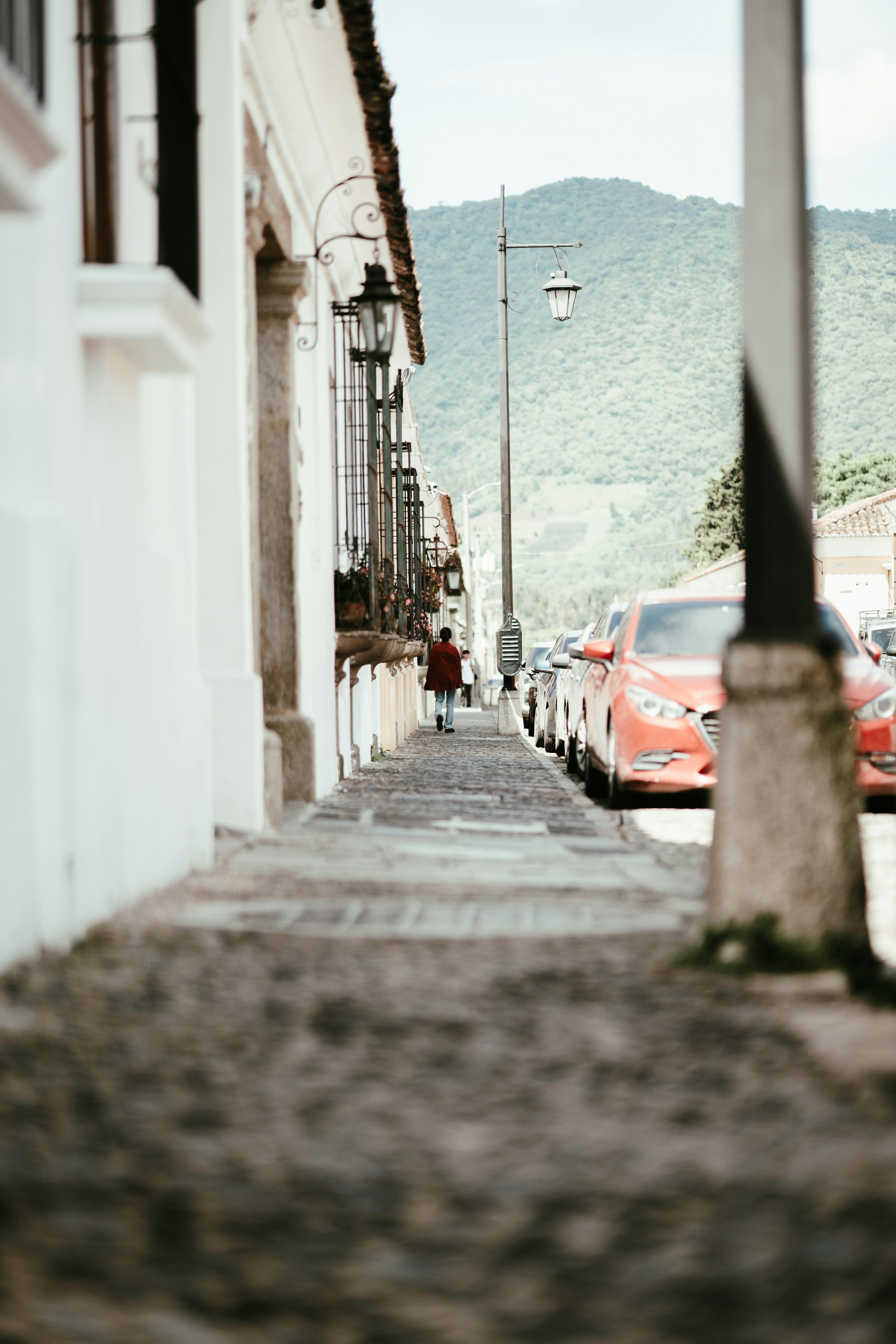 Cobblestone Corridor: A solitary walker amid car-lined facades and distant hills