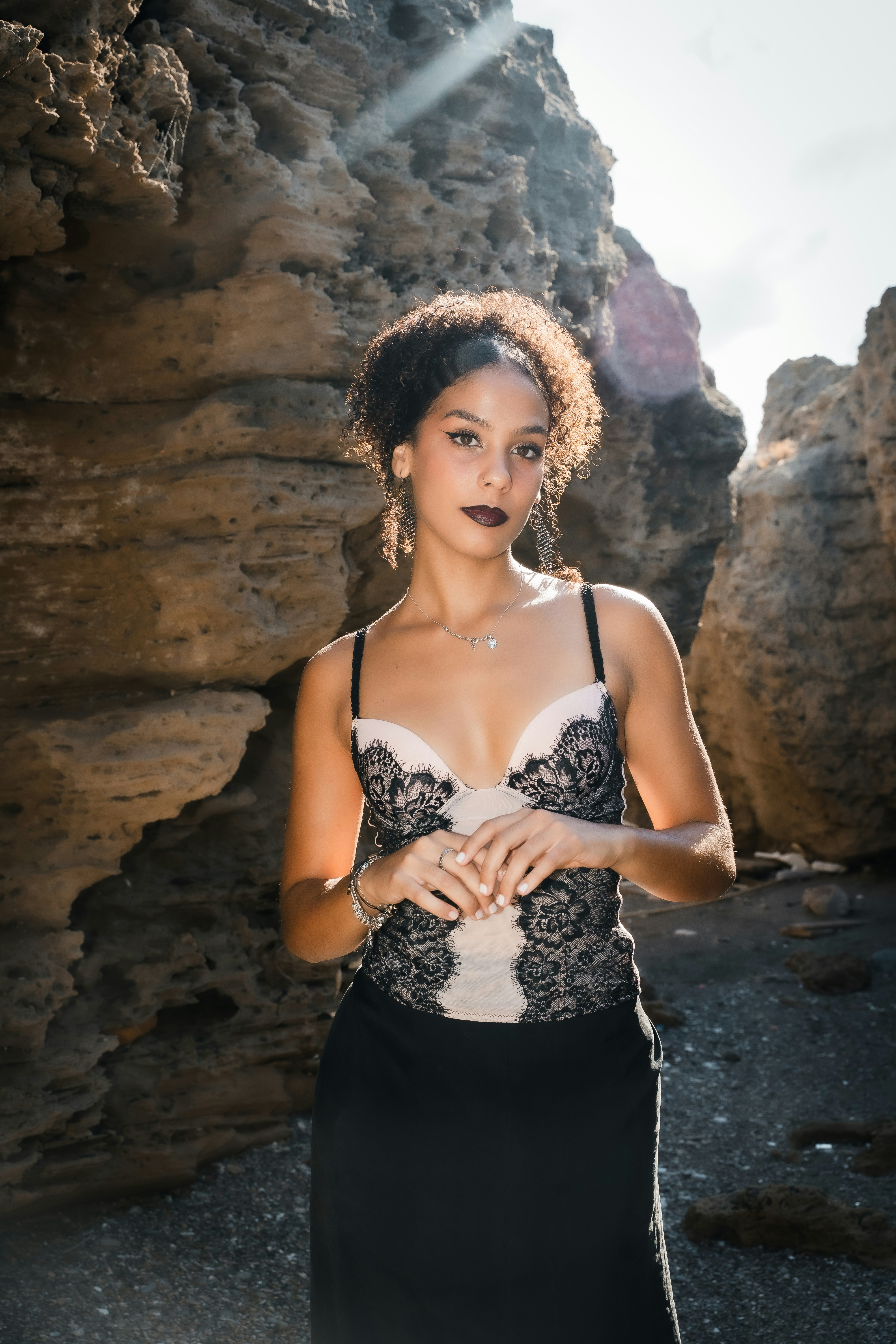 A confident woman stands against sun-lit coastal rock formations, blending elegant lace fashion with the raw textures of nature. Warm highlights illuminate her skin and curls as she poses with a calm, self-assured expression, creating a striking mix of beauty, strength, and editorial style