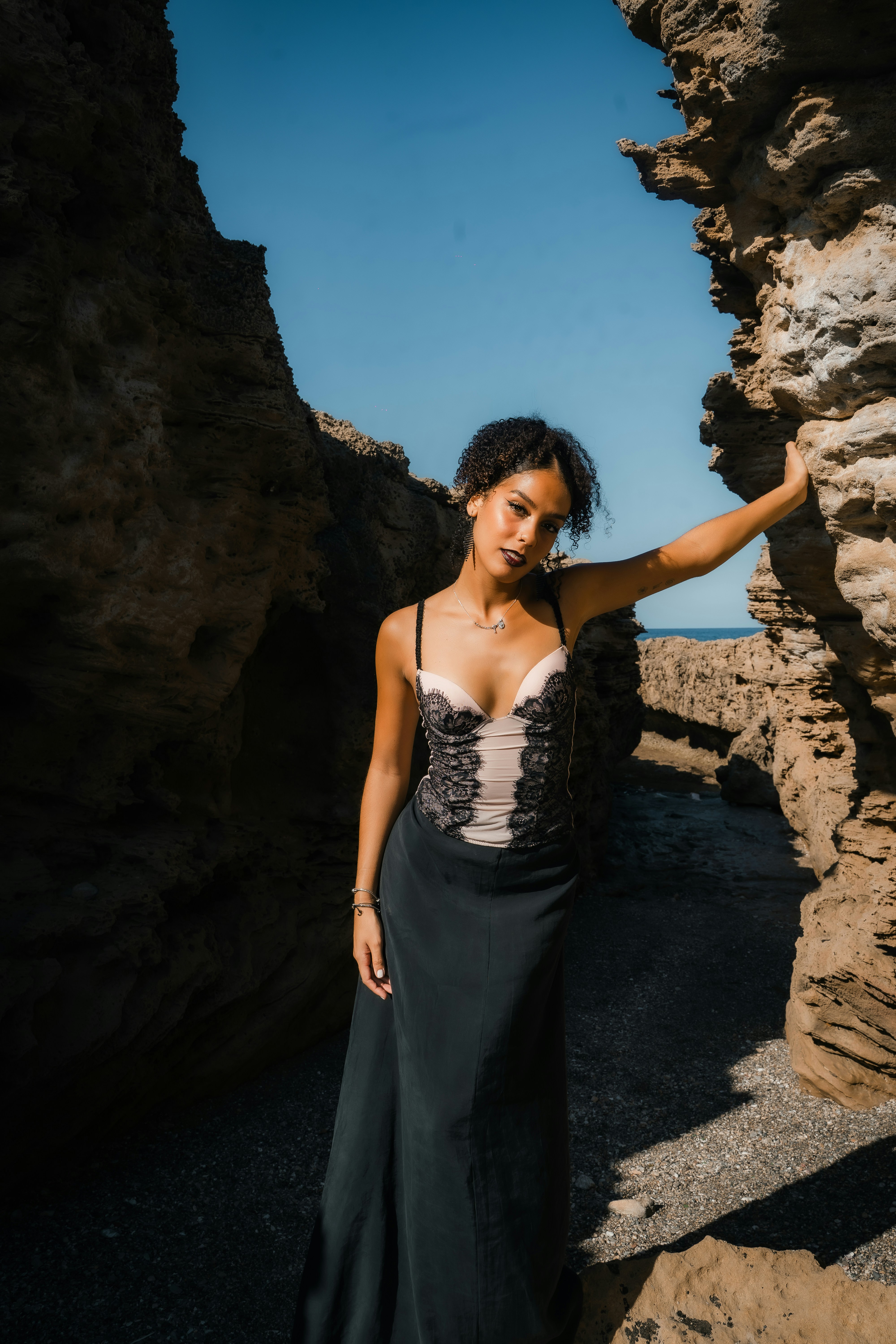 A confident woman stands against sun-lit coastal rock formations, blending elegant lace fashion with the raw textures of nature. Warm highlights illuminate her skin and curls as she poses with a calm, self-assured expression, creating a striking mix of beauty, strength, and editorial style