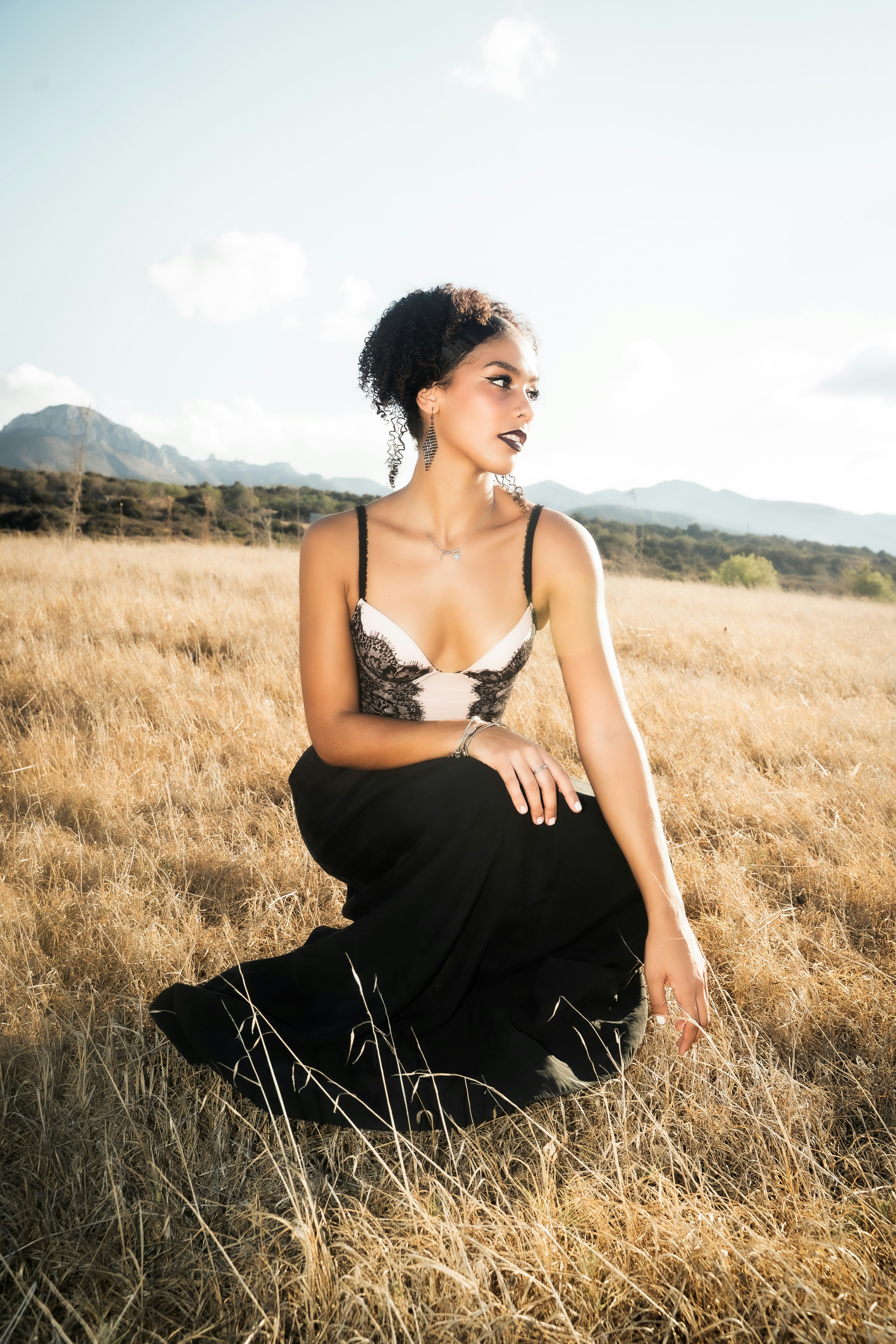 A woman stands in a golden, sunlit field with mountains in the distance, blending elegant fashion with a serene natural landscape