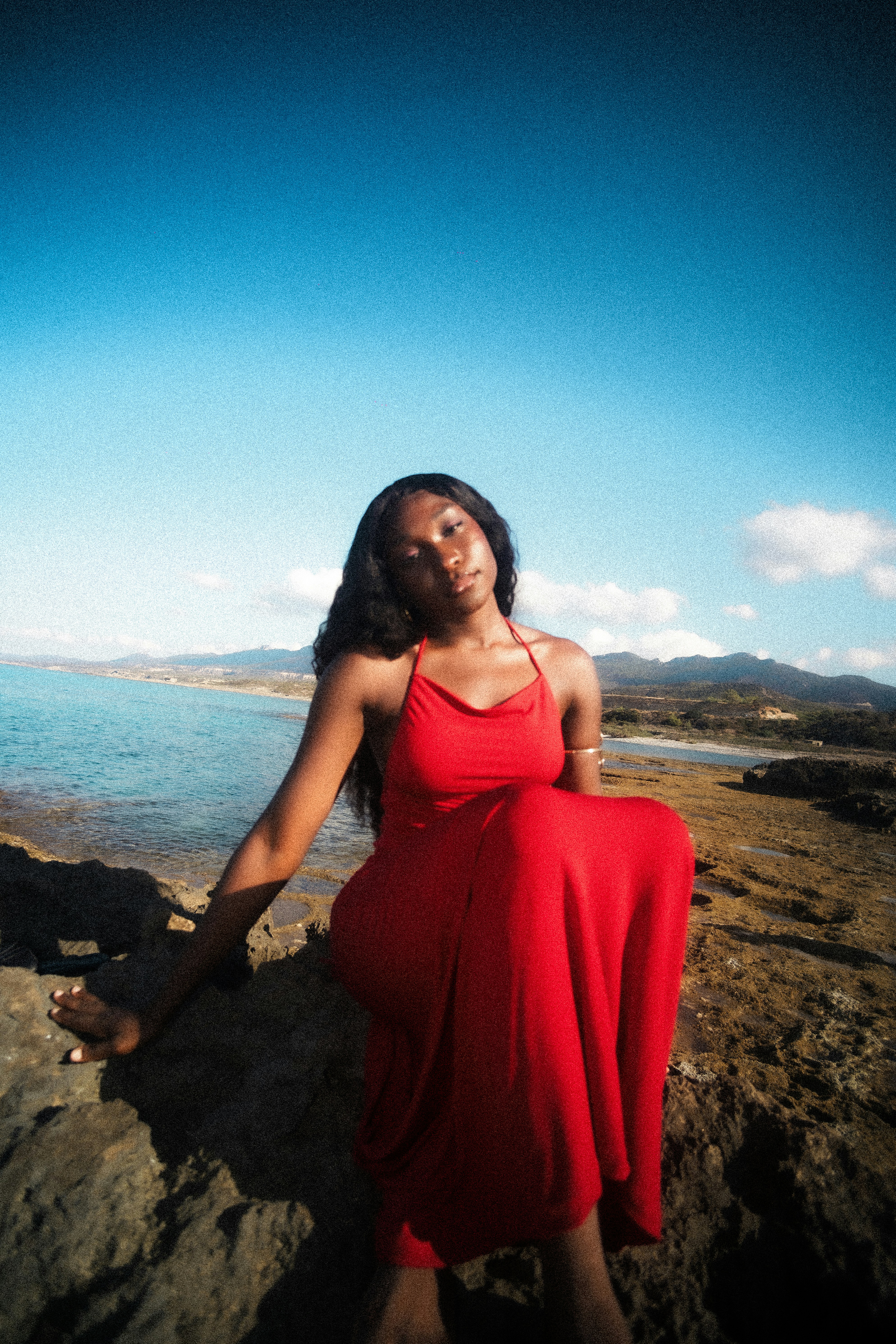 A woman in a flowing red dress reclines confidently on rugged coastal rocks, illuminated by warm sunlight. The bright blue sky and calm sea create a striking contrast with her vibrant outfit and relaxed, graceful pose.