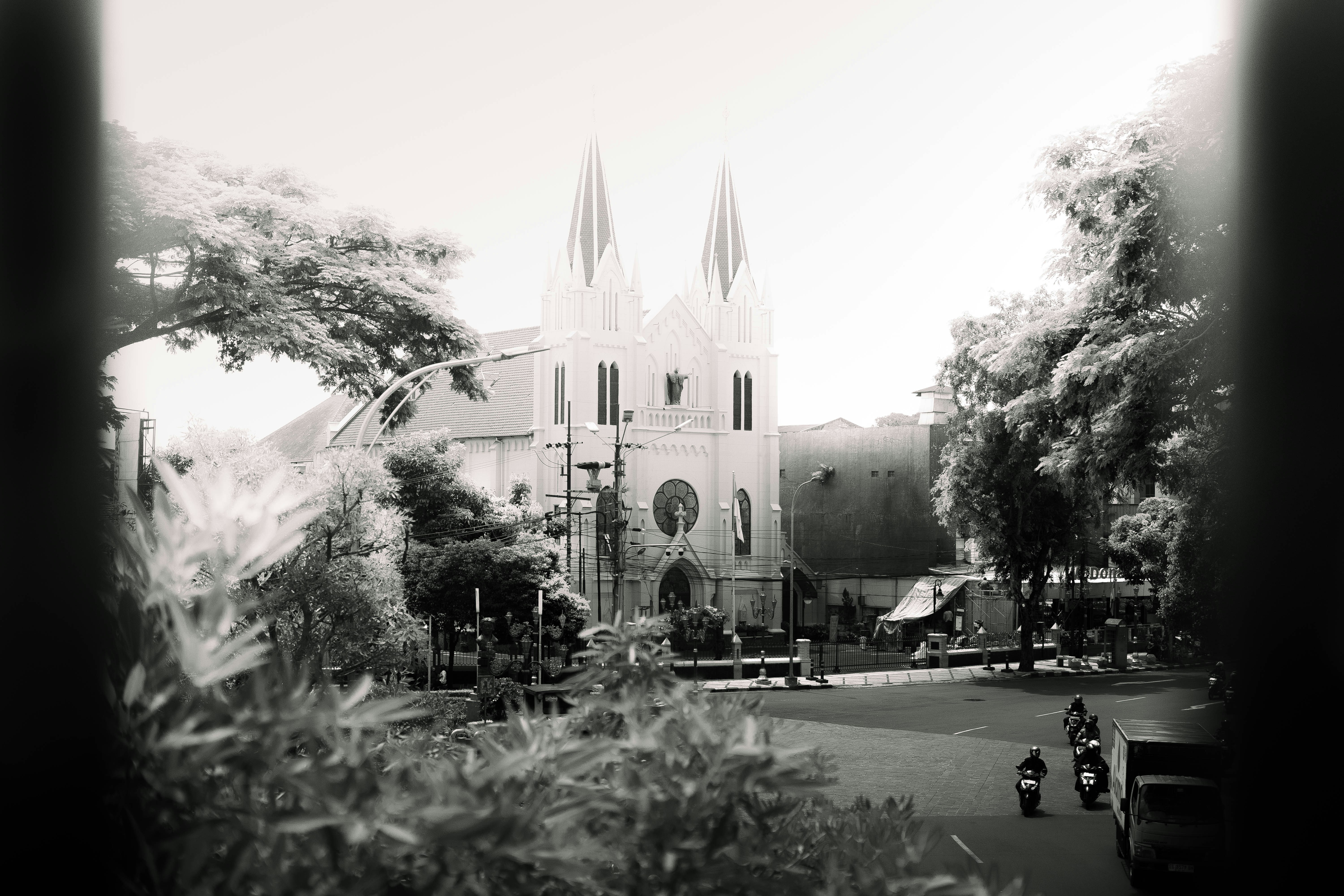 A dramatic, high-key B&W photograph of the Sacred Heart of Jesus Catholic Church in Malang, Indonesia. The extreme white light bathes the Neo-Gothic facade and the central statue of Christ in an ethereal glow, emphasizing its powerful spiritual presence. This high-contrast image is perfect for religious architecture and minimalist photography collectors.