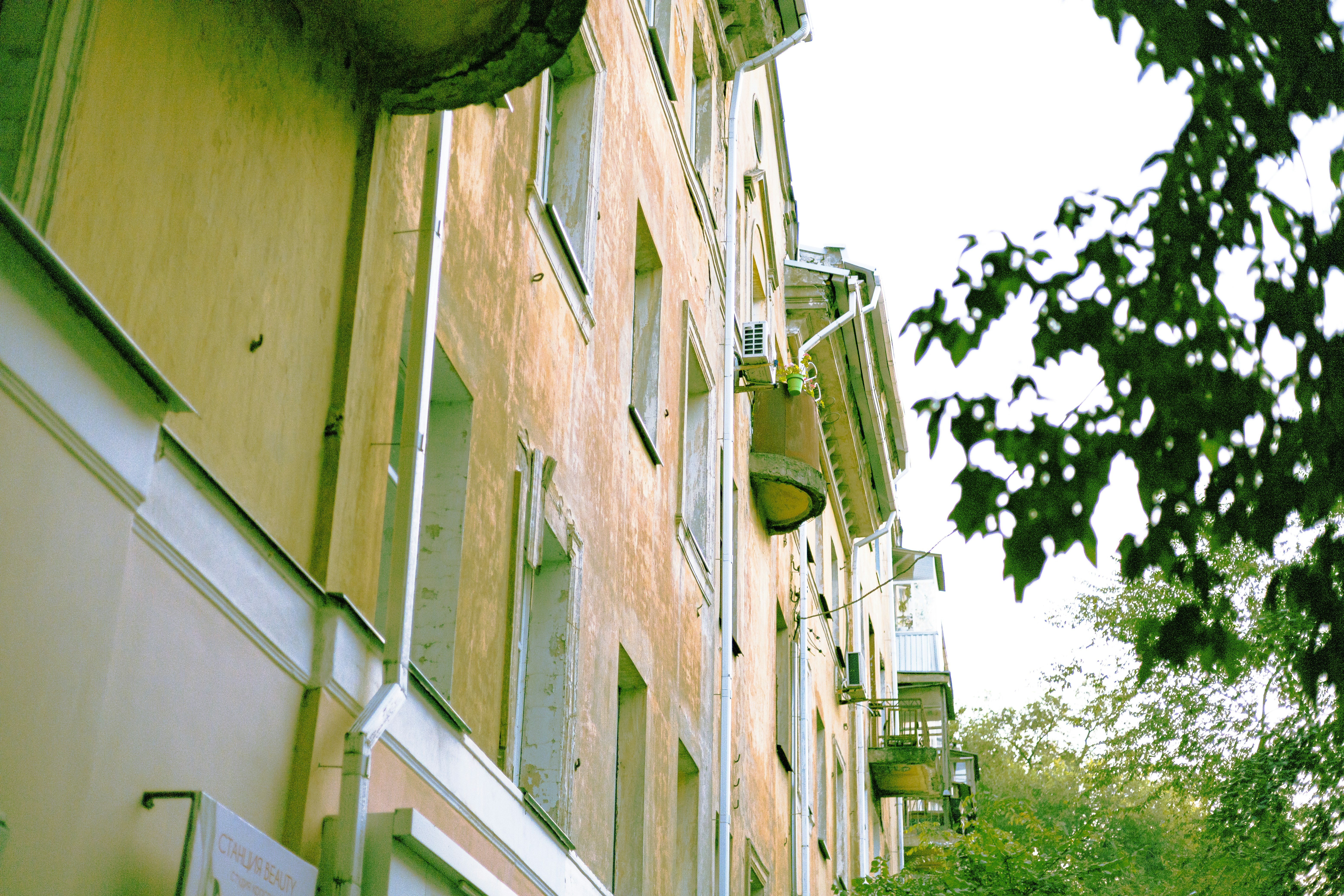 This photo captures the worn charm of an old residential building with a weathered facade, peeling paint, and small balconies that reveal its age and character. Soft daylight highlights the rustic textures, while tree branches frame the scene with natural contrast. Subtle details like air conditioners, pipes, and open windows add authenticity, making the image perfect for themes of vintage urban architecture, nostalgic city life, and atmospheric streetscapes.