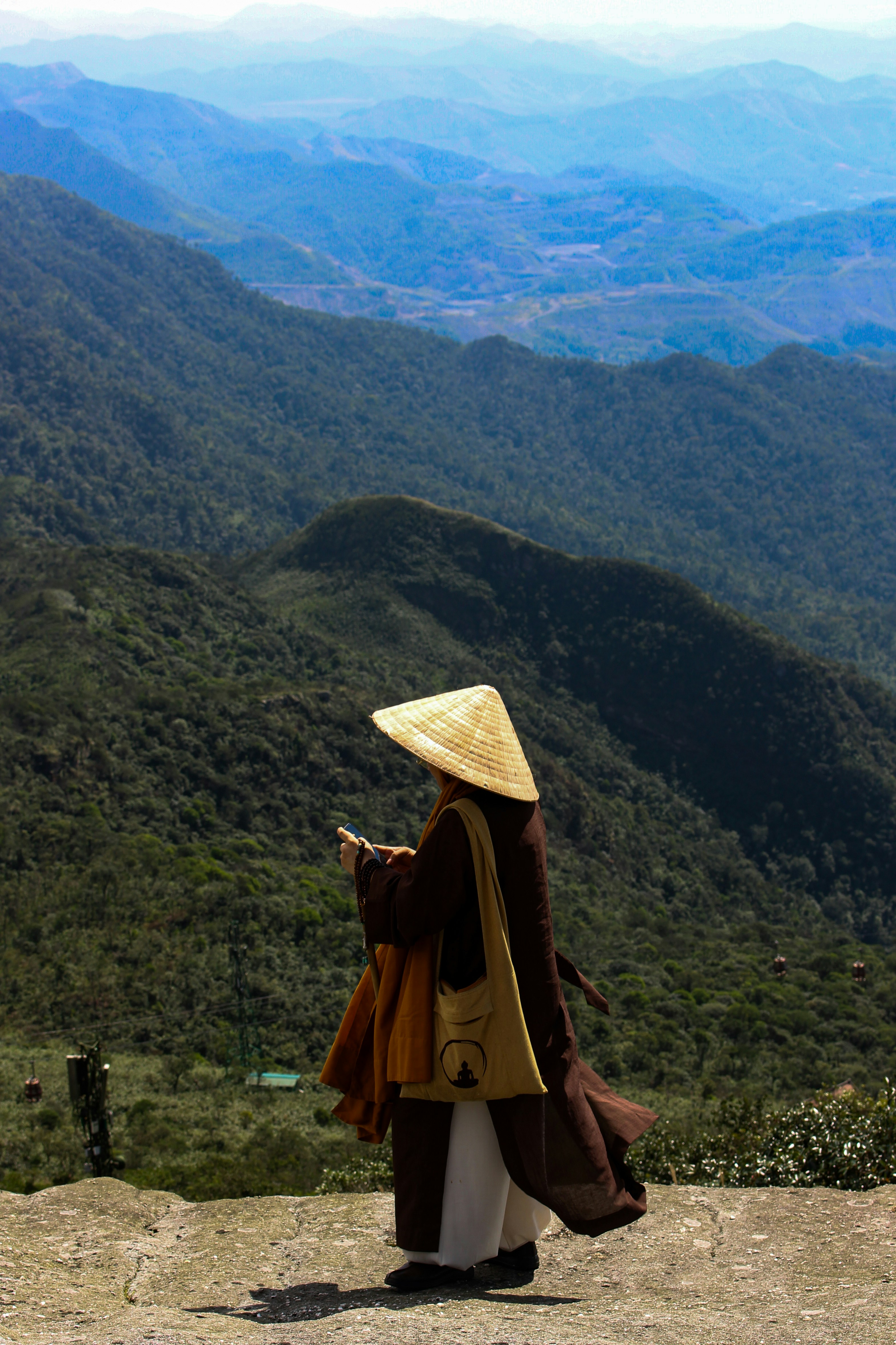 Photograph of a traveler in a conical hat standing on a terrace, gazing toward layered mountain ranges.