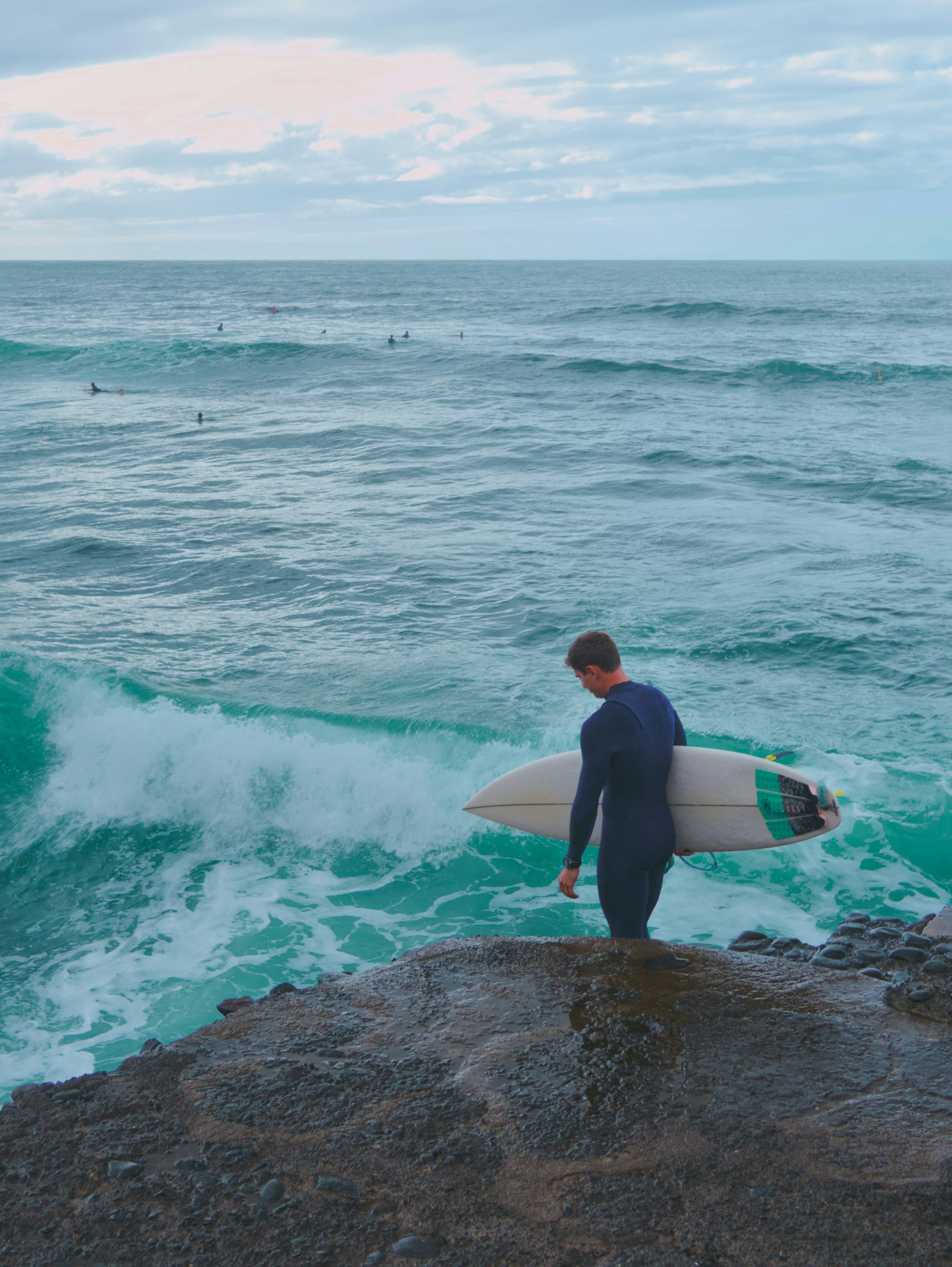 Edge of the Break: A lone surfer strides along rugged rocks toward emerald swells