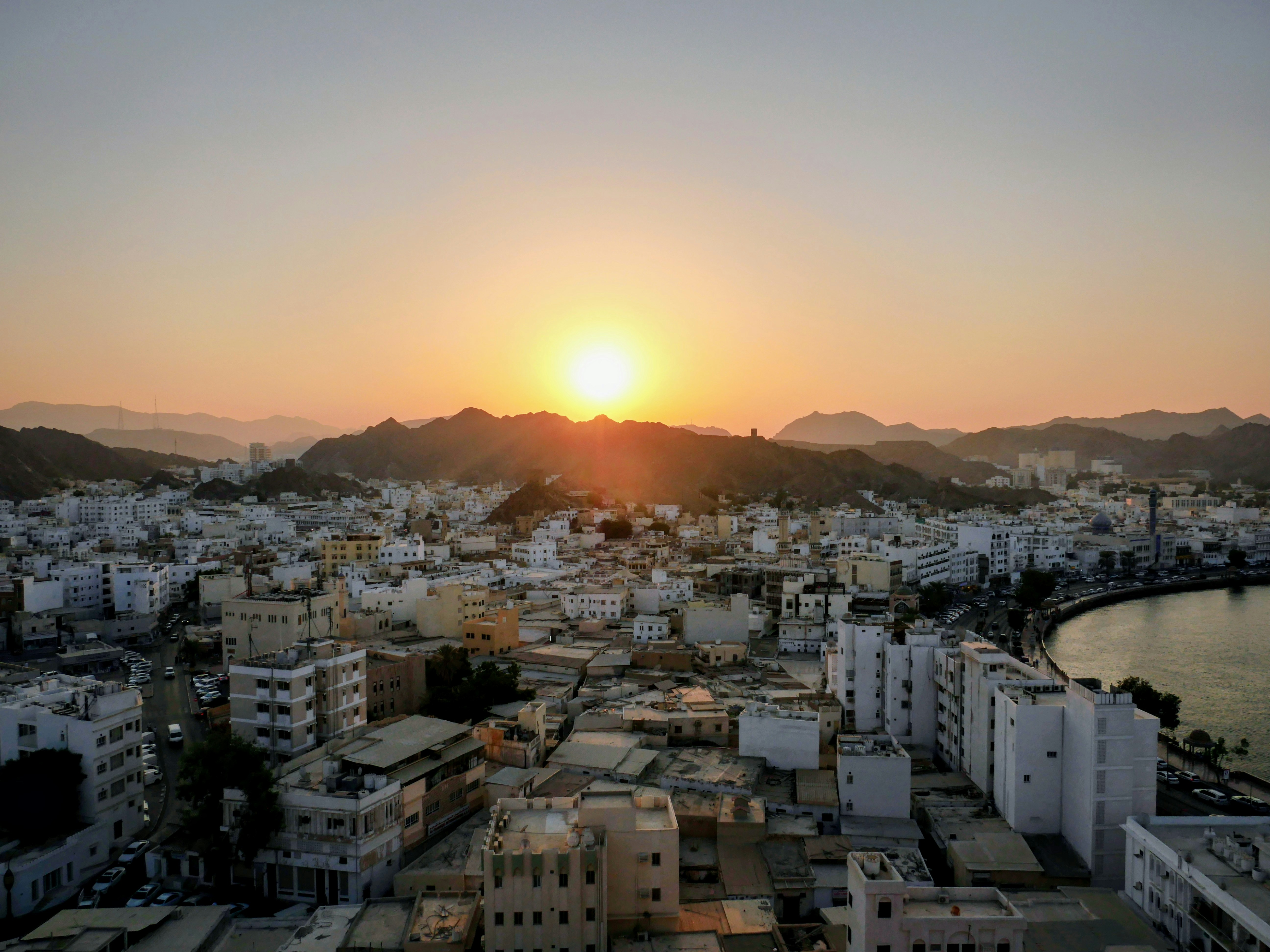 A panoramic view of Muttrah, Muscat’s historic port district, bathed in the soft light of dusk. The image captures the bustling harbor, traditional Omani buildings, and the rugged coastline, reflecting the blend of maritime heritage and urban charm. The warm tones of the setting sun add a magical touch to this coastal cityscape.