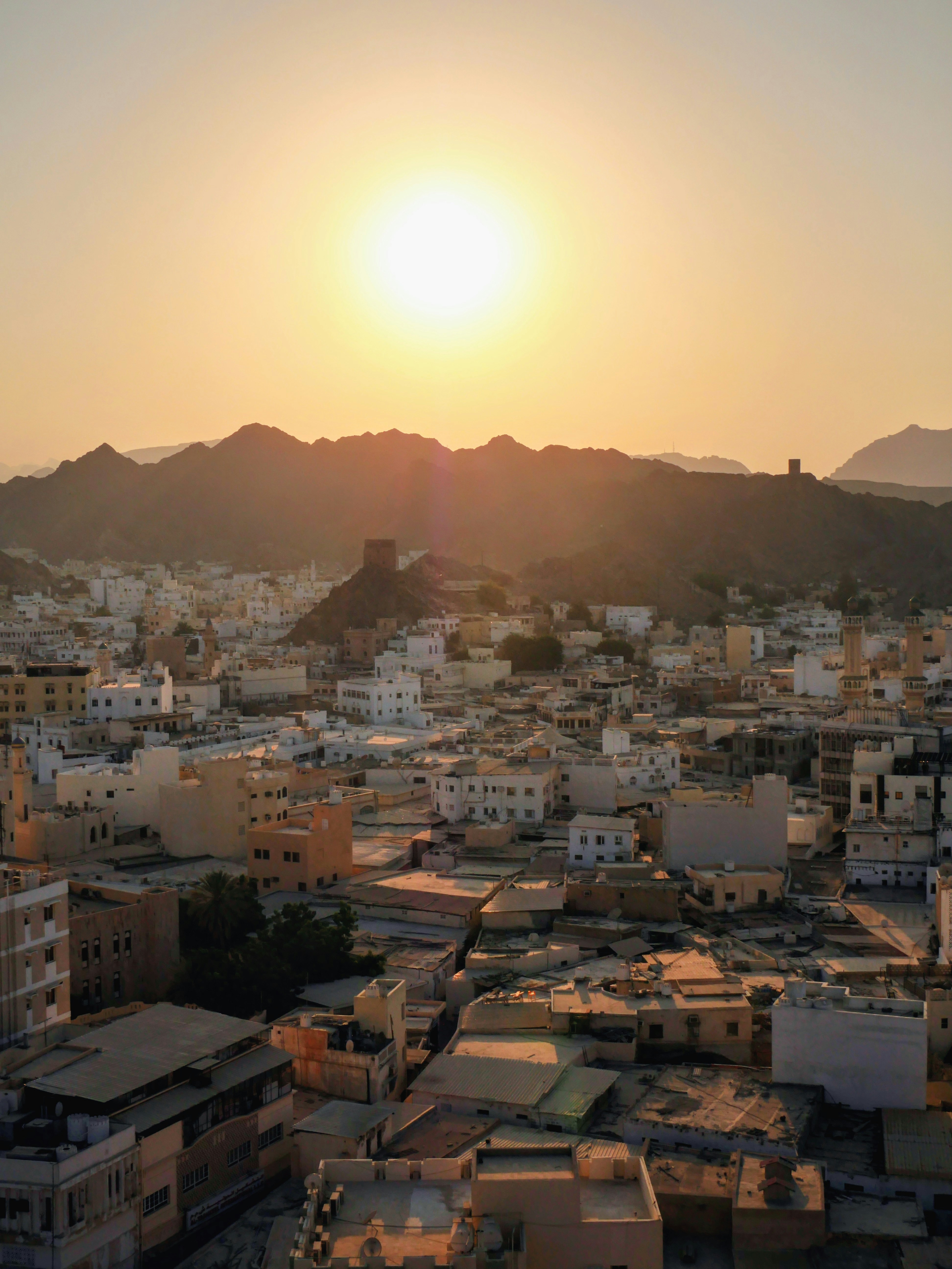 A stunning sunset over the historic city of Muscat, Oman, casting a warm golden glow across the urban landscape. The sun dips behind the rugged mountains, illuminating the city with soft, vibrant hues. This image captures the serene beauty of Oman’s capital, blending tradition and modernity under a breathtaking sky.