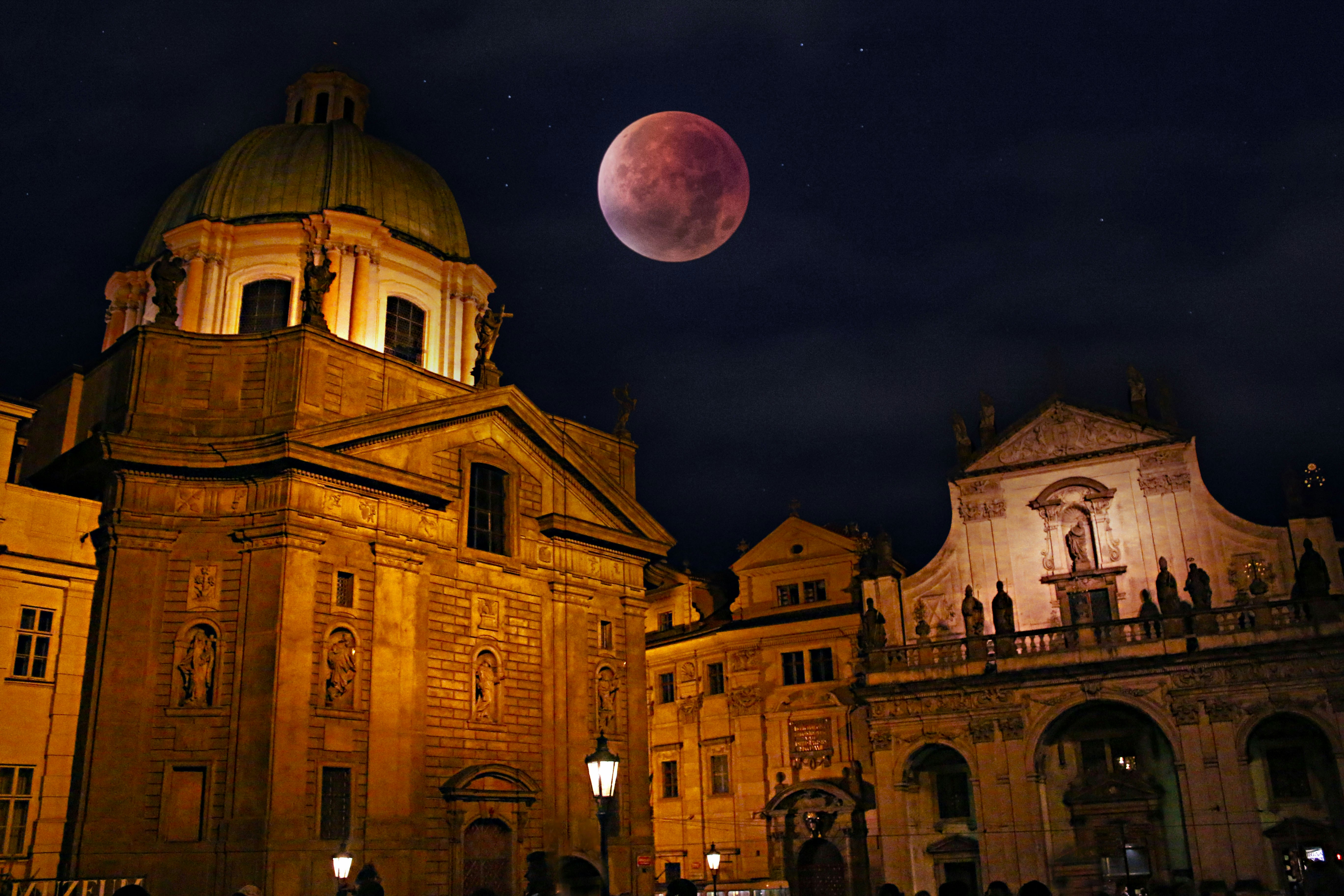 La Plaza Křižovnické, situada junto al inicio del Puente de Carlos, se ilumina con la magia de un eclipse lunar. La cúpula de la iglesia de San Salvador y la fachada barroca de la iglesia de San Francisco de Asís enmarcan la escena, donde la luna roja añade un toque místico al corazón histórico de Praga.