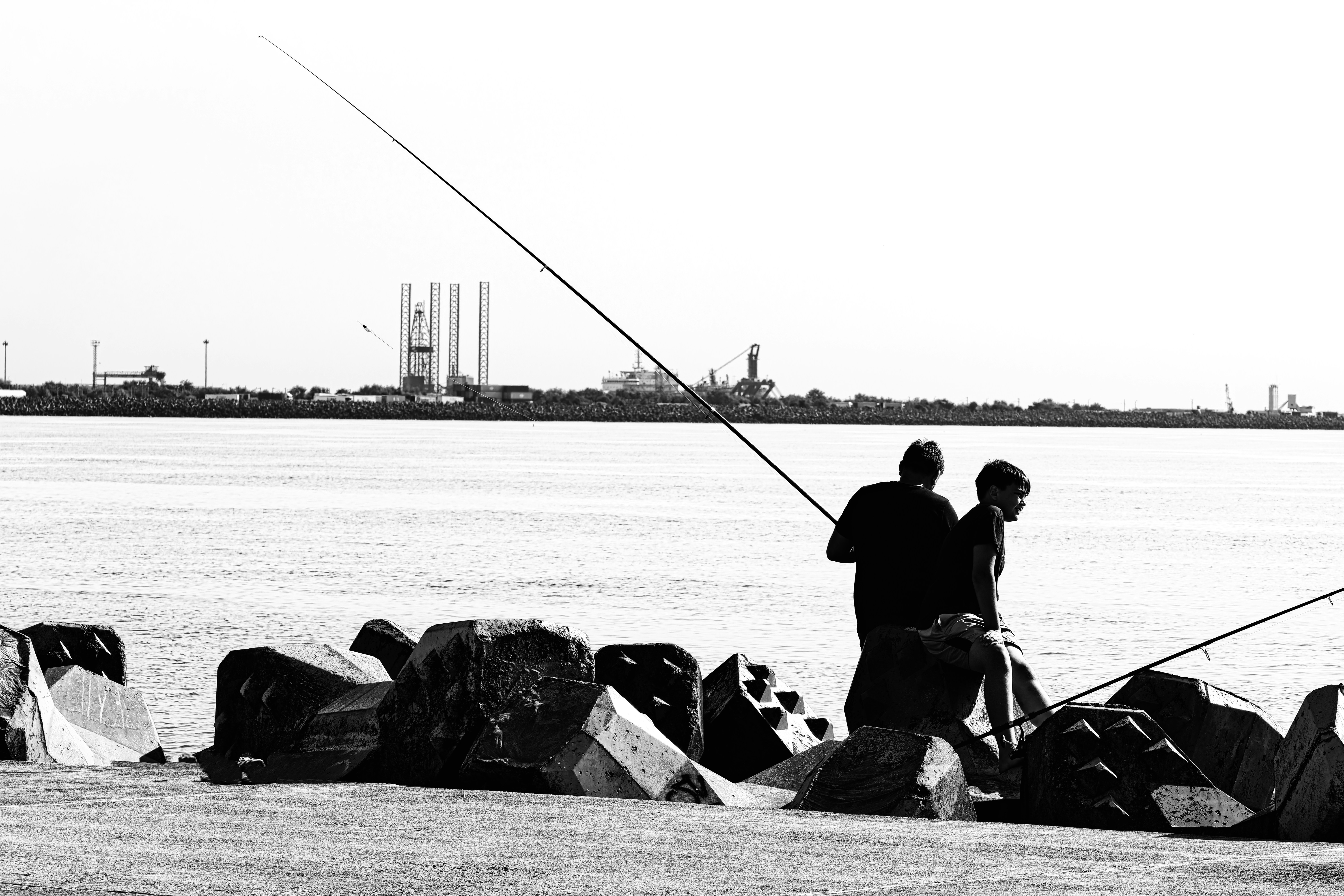 Breakwater Silhouettes: Fishing Duo Against a Distant Industrial Horizon