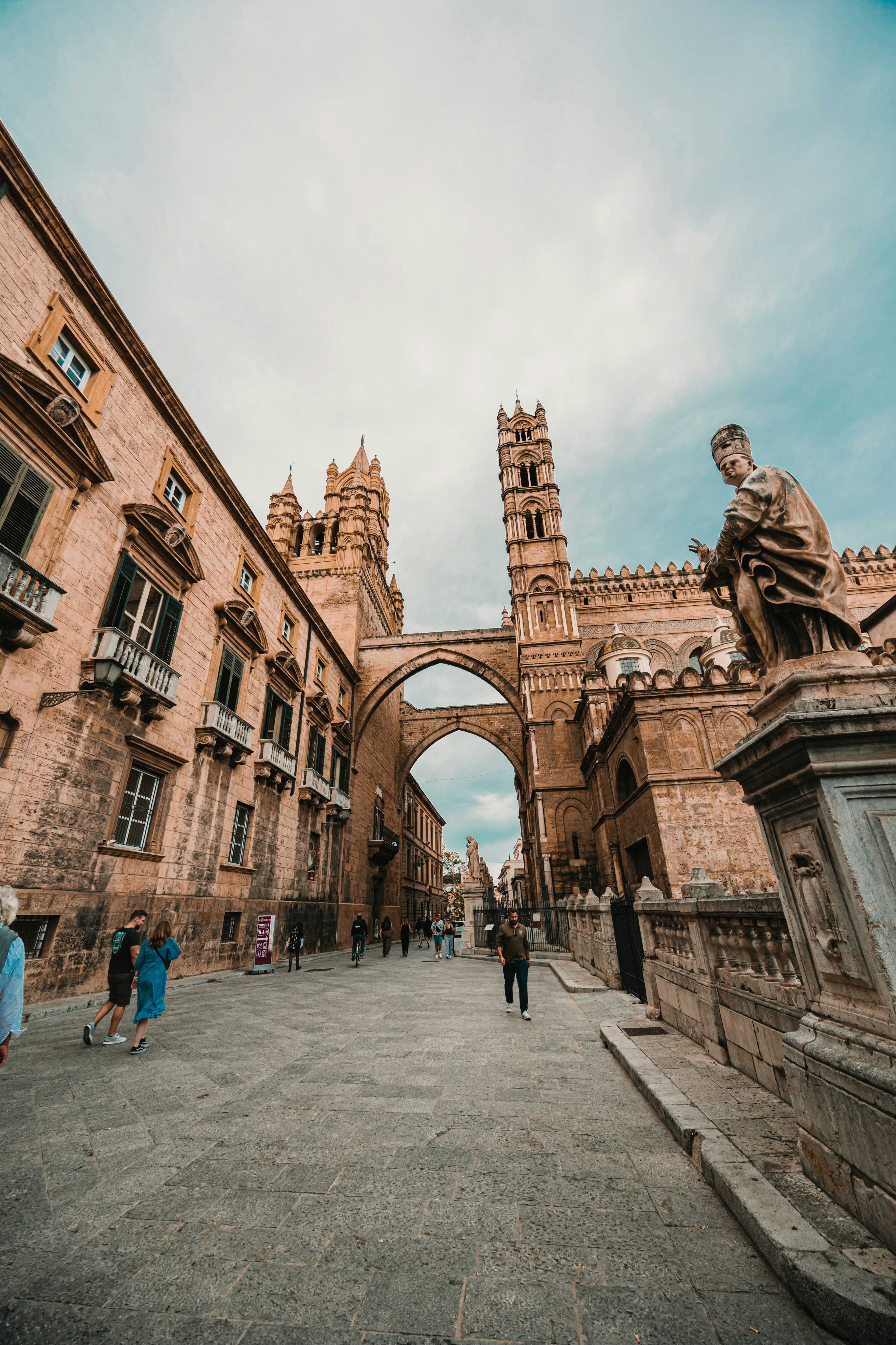 Street view of Palermo Cathedral's entrance and bell tower, showing the arched walkway over the street and pedestrians in the foreground.