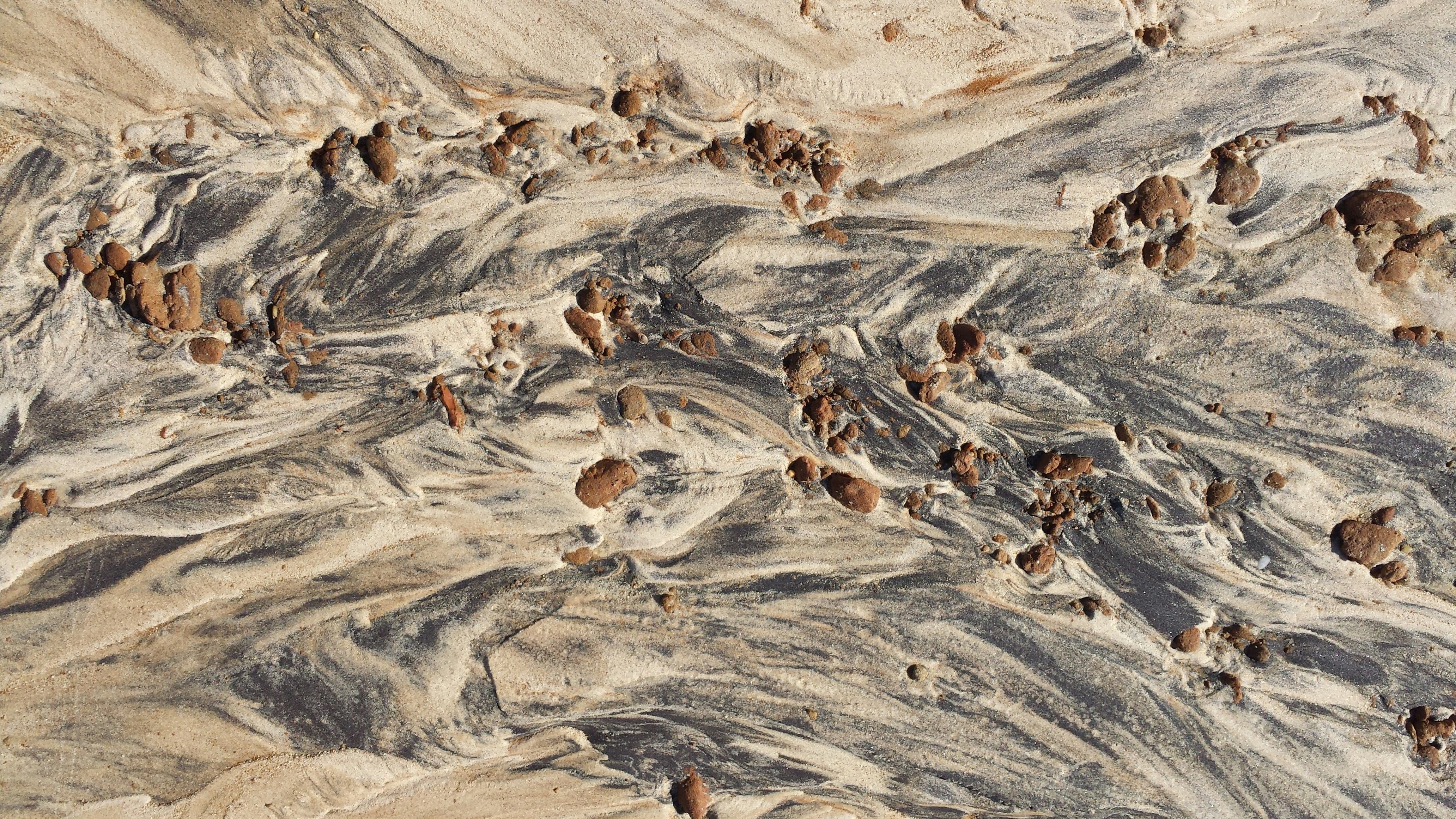 A close-up of wet sand, featuring patterns created by water flow. The lighter areas are sand, while the darker, vertical streaks are heavy mineral deposits concentrated by the action of water. The small, round, reddish-brown clumps are mineral concretions or perhaps small pebbles which might contain iron-oxide due to the color.
