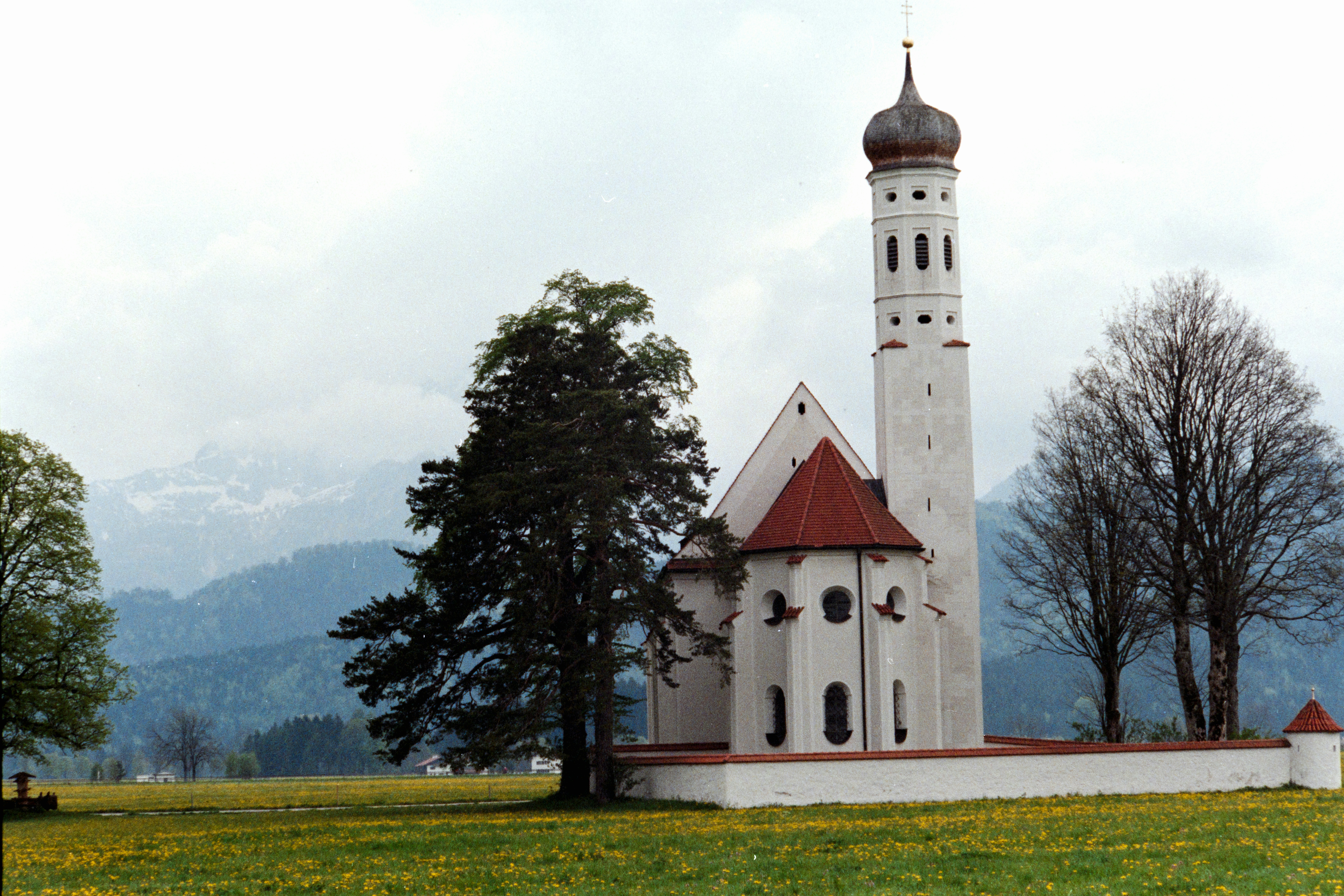 White Chapel and Red-Tiled Tower Rising Over Meadow Toward Distant Mountains