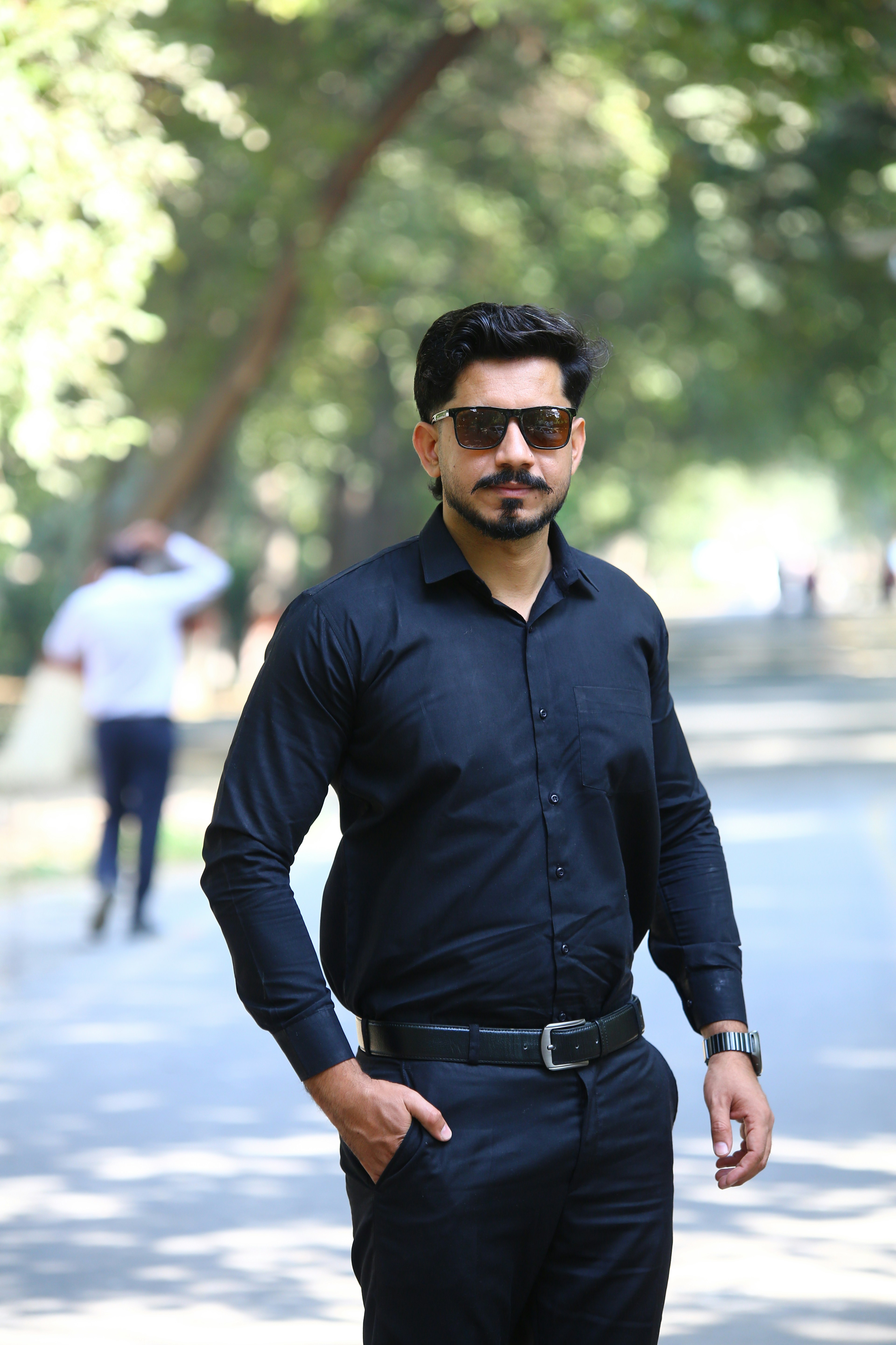 A stylish street-style portrait featuring black pants and shirt, sunglasses, and phone, captured at Kanan Park, Sahiwal. Perfect for wedding, event, or lifestyle-related content. Credit appreciated: ShadiCenter.com for providing this image.