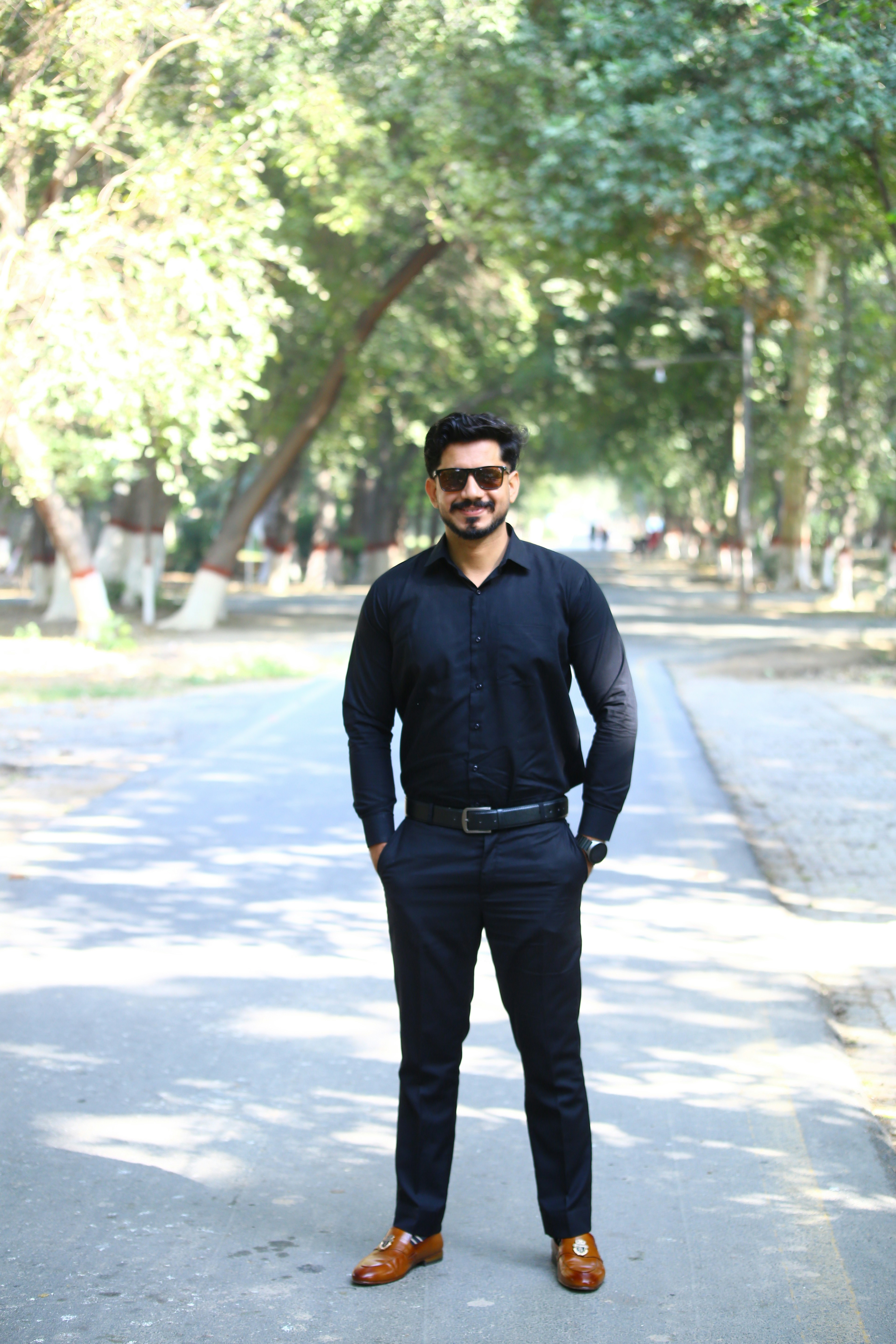 A stylish street-style portrait featuring black pants and shirt, sunglasses, and phone, captured at Kanan Park, Sahiwal. Perfect for wedding, event, or lifestyle-related content. Credit appreciated: ShadiCenter.com for providing this image.