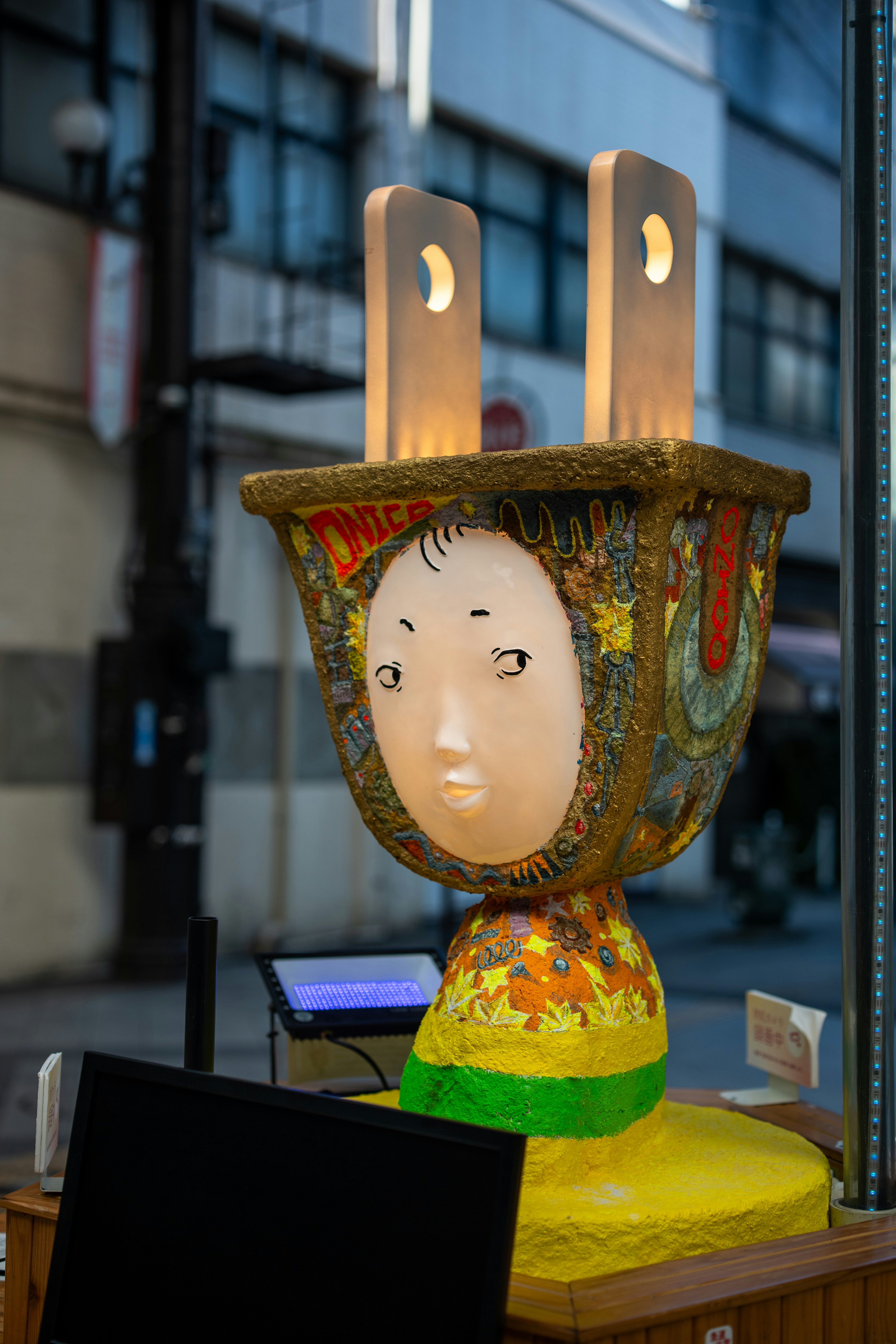 Face-Bust in a Window: A Colorful Ceramic Sculpture Under Warm City Lights