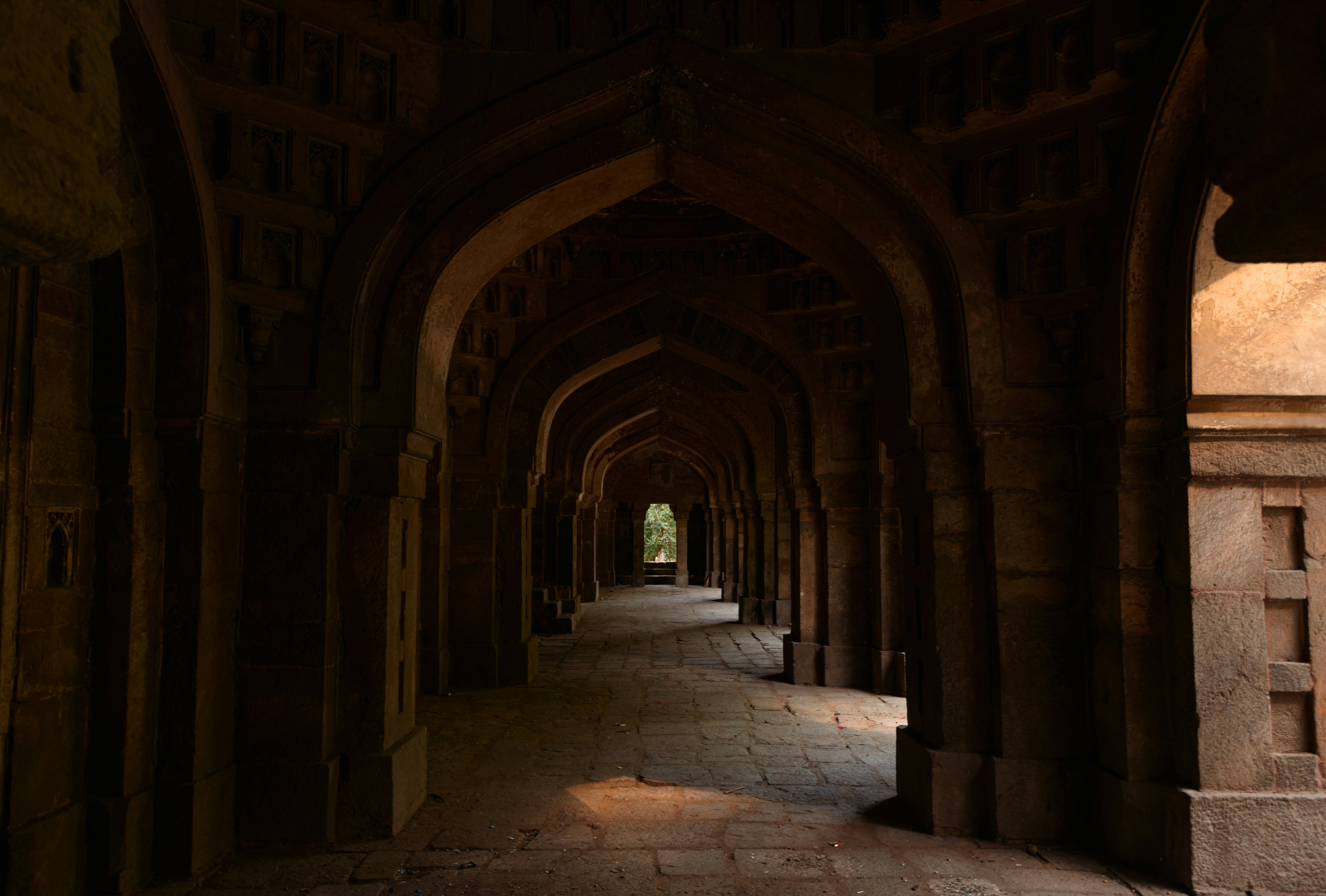 The interior corridor of Masjid Moth is built entirely of stone and lined with repeating arches through which warm light filters in. This mosque dates to the Lodhi era and was commissioned in the early 1500s. According to historical accounts, Sultan Sikandar Lodi granted a bag of moth lentils to his prime minister, Wazir Miya Bhoiya, who used its sale to fund the construction of the mosque. Over time the structure became known as Masjid Moth in reference to that origin. The building reflects characteristic Lodhi period work with robust stonework, pointed arches, and sequences of vaulted bays.