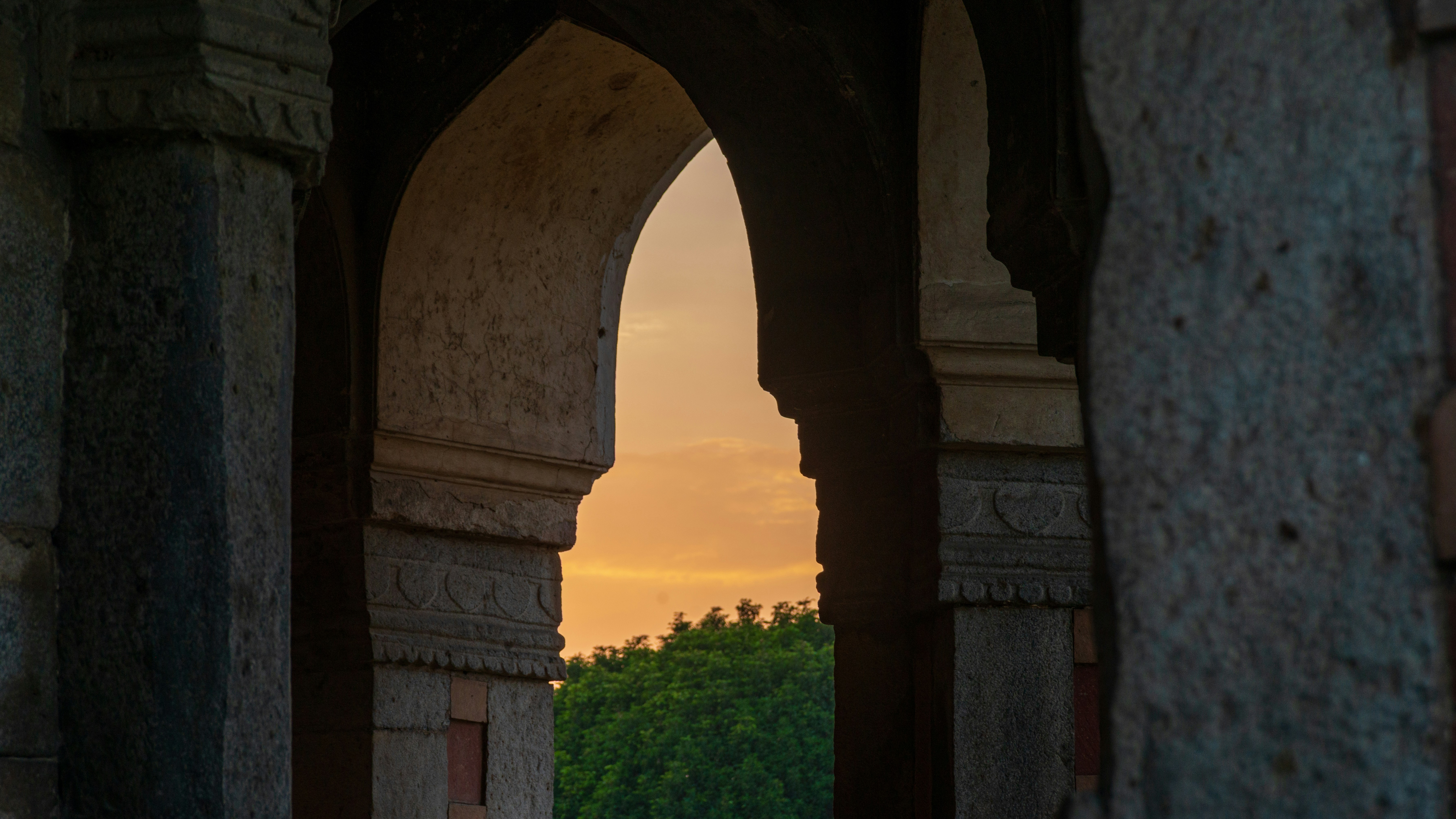 A quiet moment inside the stone corridors of Isa Khans Tomb in Delhi. The pointed arch frames a glowing sunset as warm light contrasts with the cool textured stone of the monument. The silhouette of the arch opens toward dense green trees outside creating a layered view that blends Lodhi era architecture with the calm of nature. The scene captures the peaceful atmosphere of this historic enclosure and the refined geometry of Indo Islamic design.