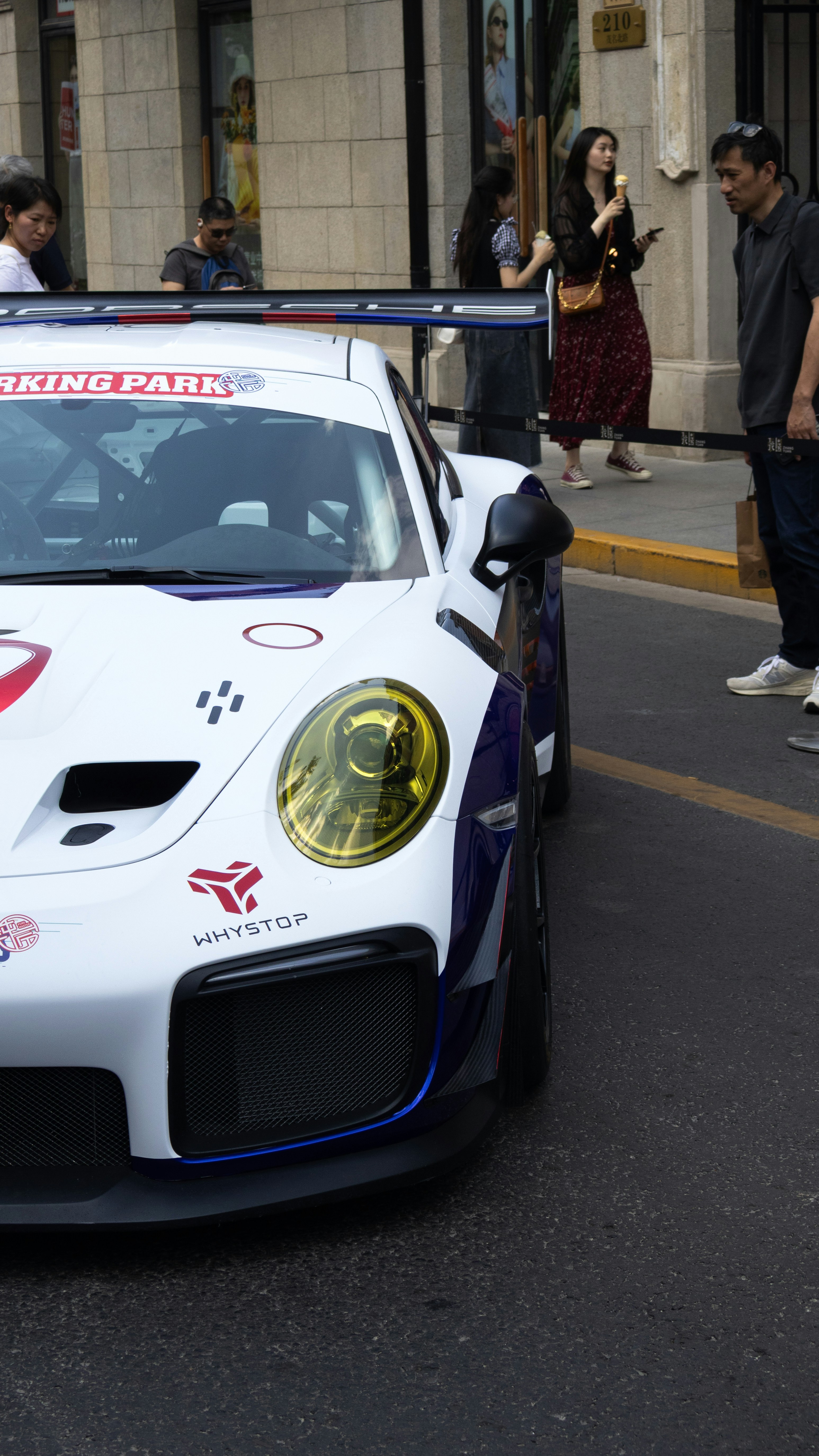 A white Porsche race car with yellow headlights parked on a city street in Shanghai while people walk past. The racing livery, large rear wing and WHYSTOP logo contrast with the everyday urban scene, blending motorsport culture with city life.