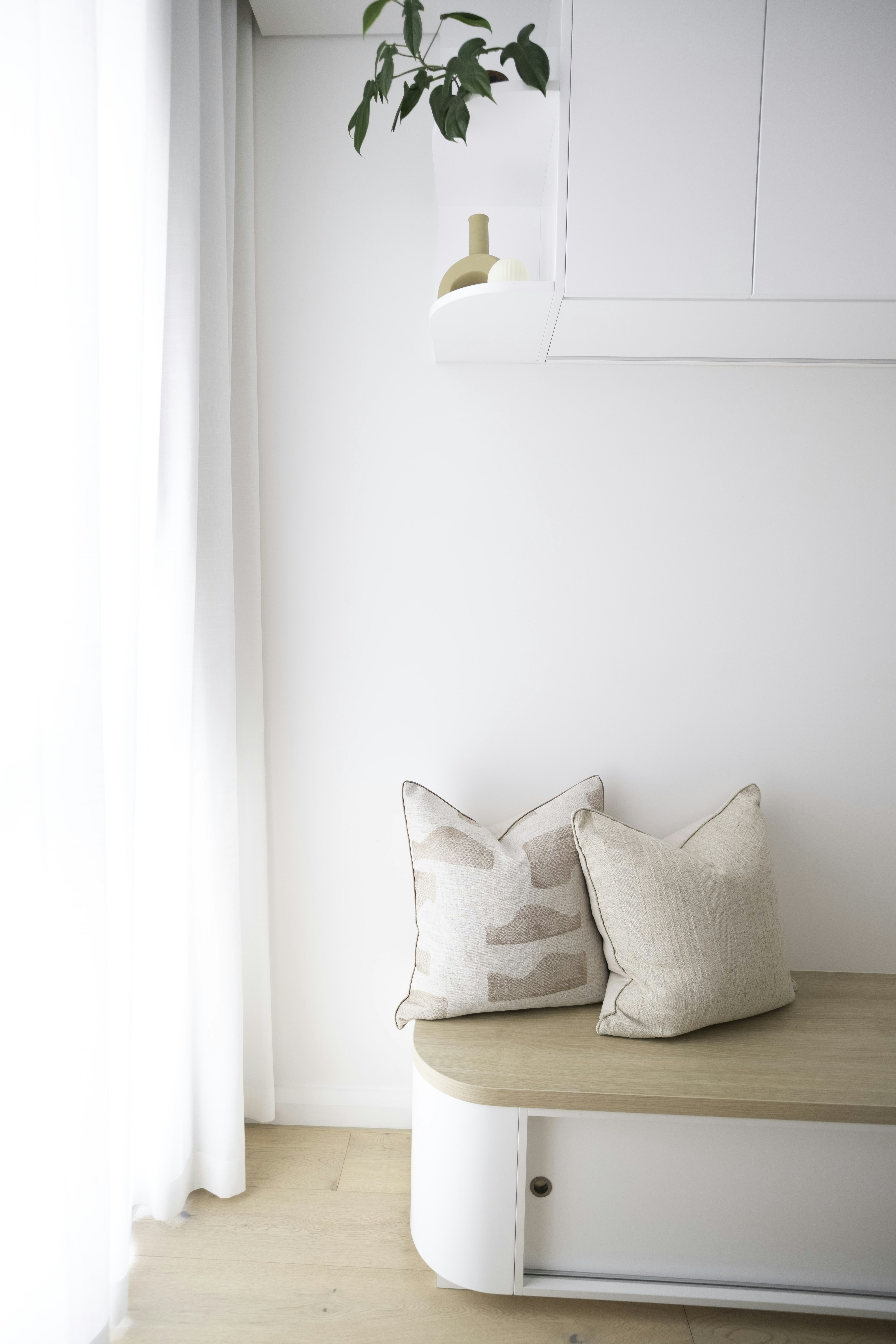 A soft, minimalist corner with two textured neutral cushions styled on a light timber bench. Sheer curtains filter gentle daylight into the space, creating a calm and airy interior. A simple sculptural vase and green foliage add a natural touch, enhancing the serene, Japandi-inspired atmosphere.