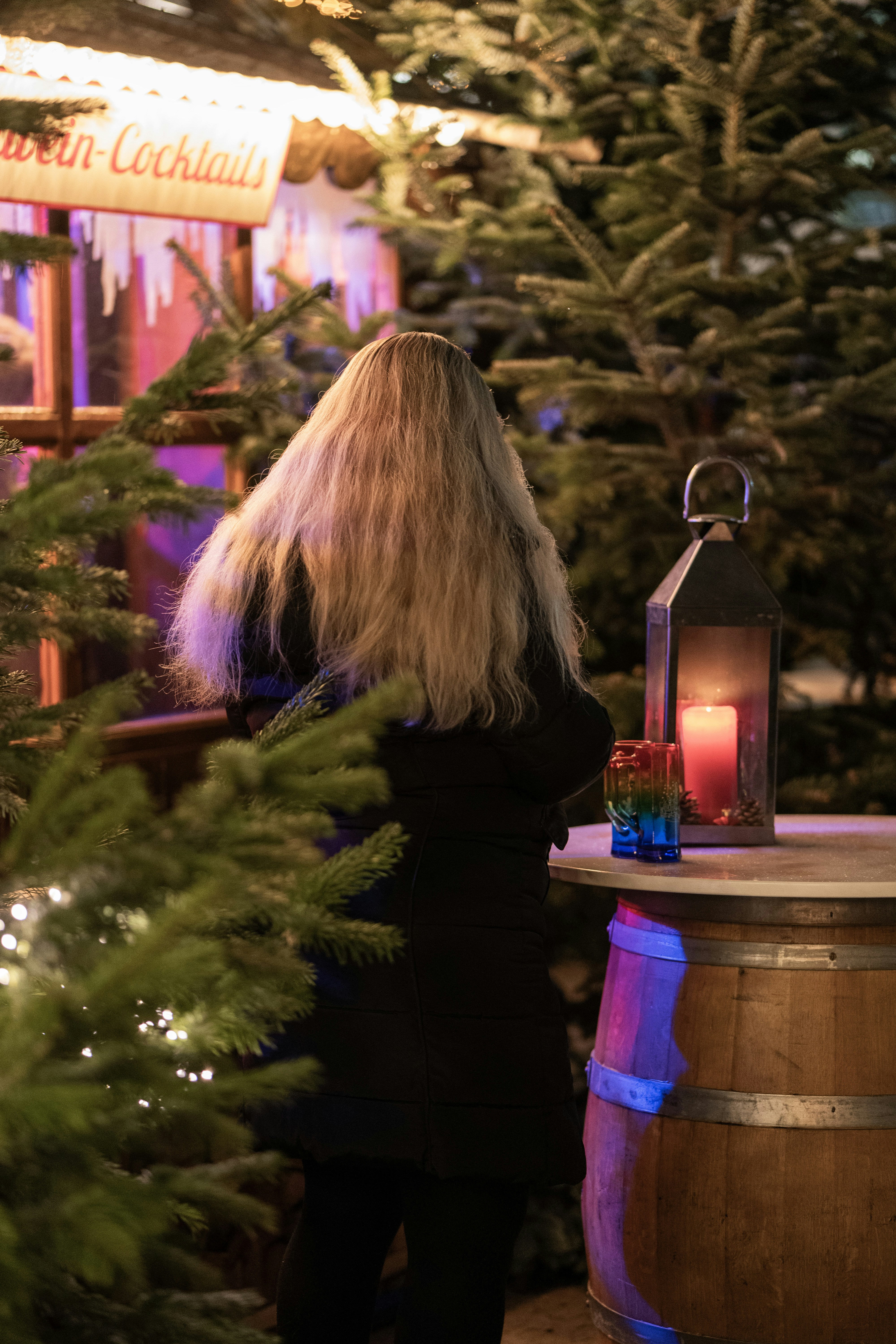 Frau mit langen blonden Haaren steht auf dem Weihnachtsmarkt an einem Stehtisch mit Kerze umgeben von Tannen.