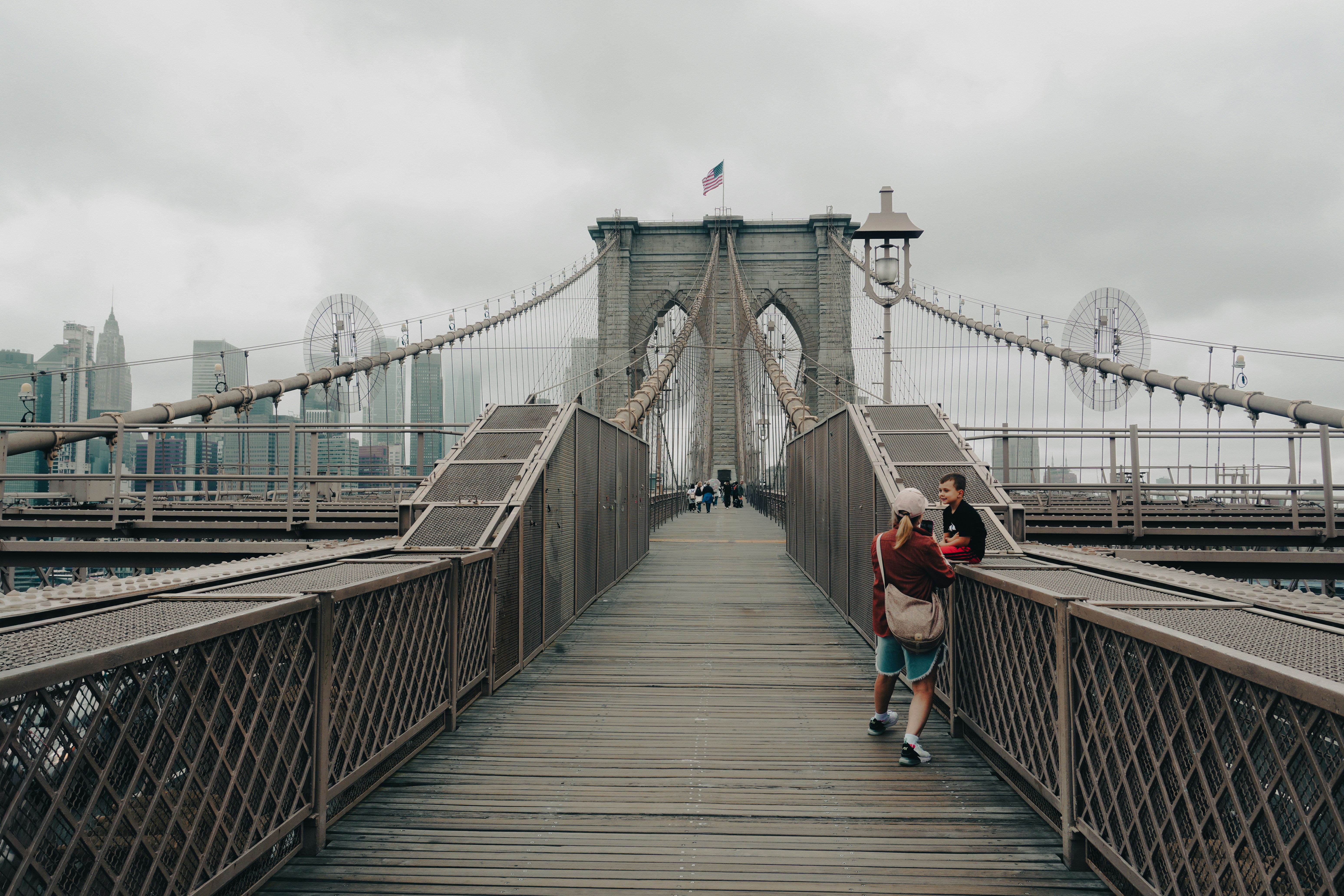 A wide-angle view of the Brooklyn Bridge walkway on a cloudy day, showcasing its iconic architectural cables, stone towers, and the New York City skyline in the background. A calm urban moment capturing tourists and the timeless beauty of one of NYC’s most famous landmarks.