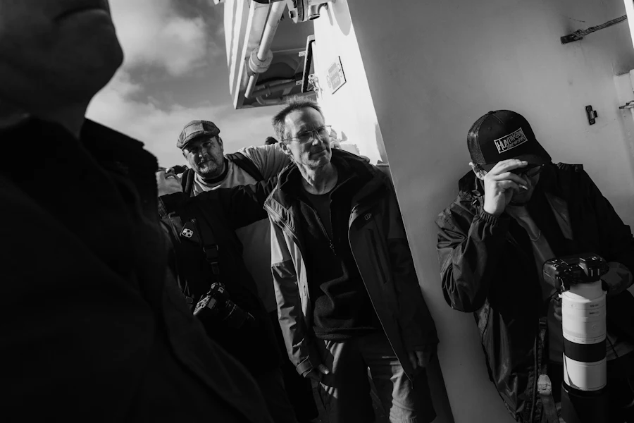 Black-and-white candid scene of three men standing on a boat or ship deck, with one adjusting his cap beside a mounted post in the foreground. A large wall and cloudy sky frame the group.