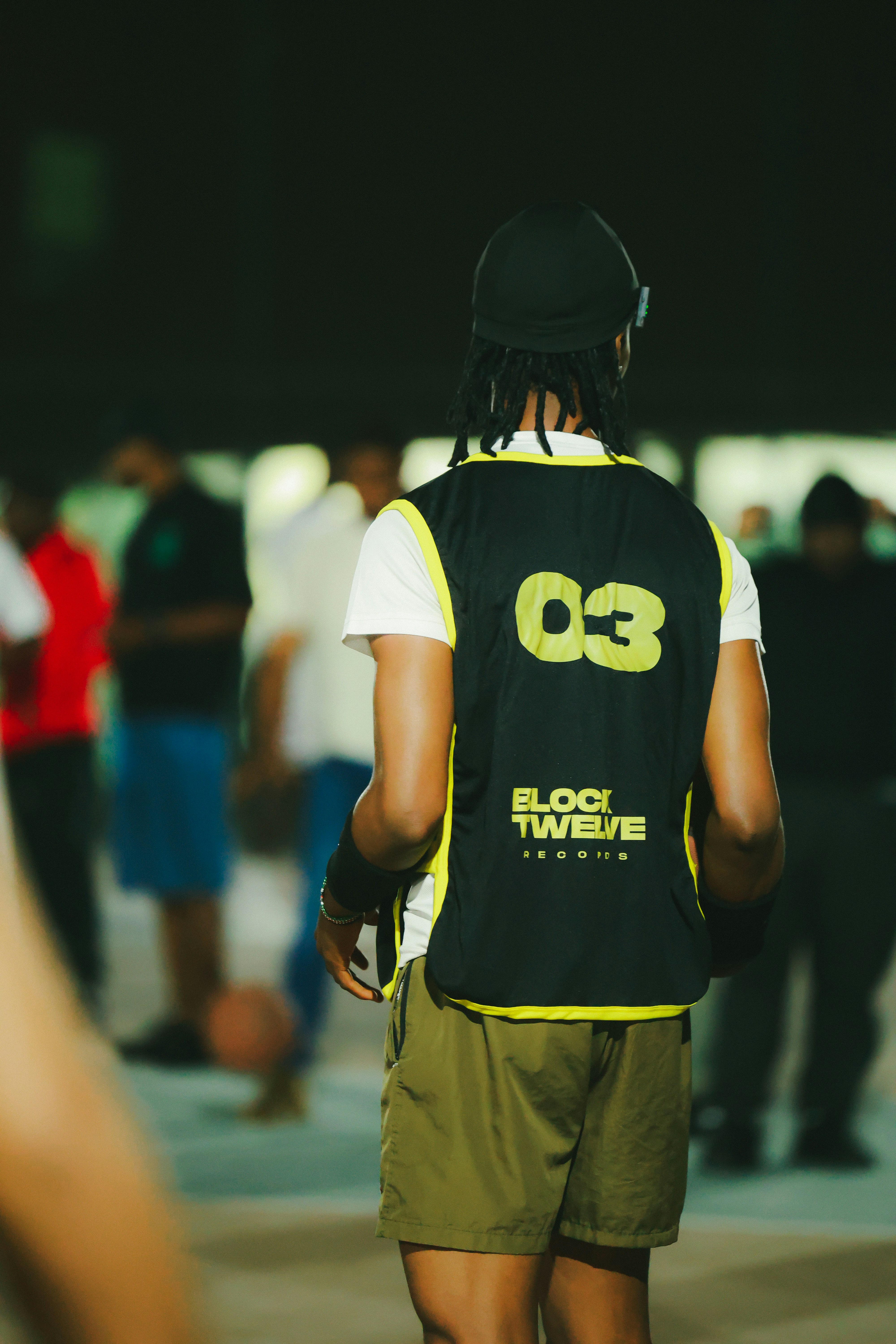 Back of a person in a black jersey with yellow trim and the number 03 stands among a blurred crowd in an indoor setting.