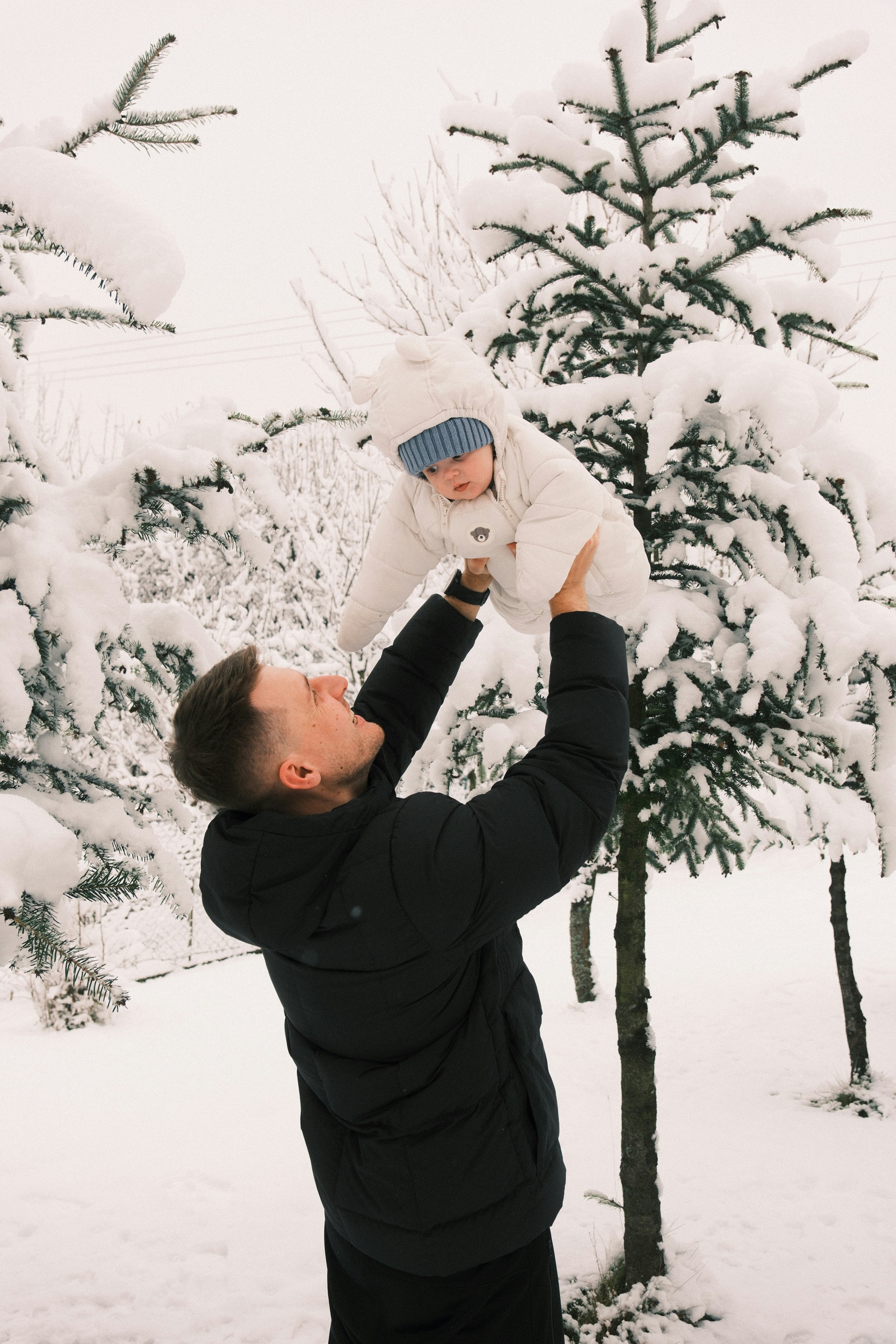 Snow-Kissed Lift: Father Lifts Baby Among Frost-Blanketed Pines