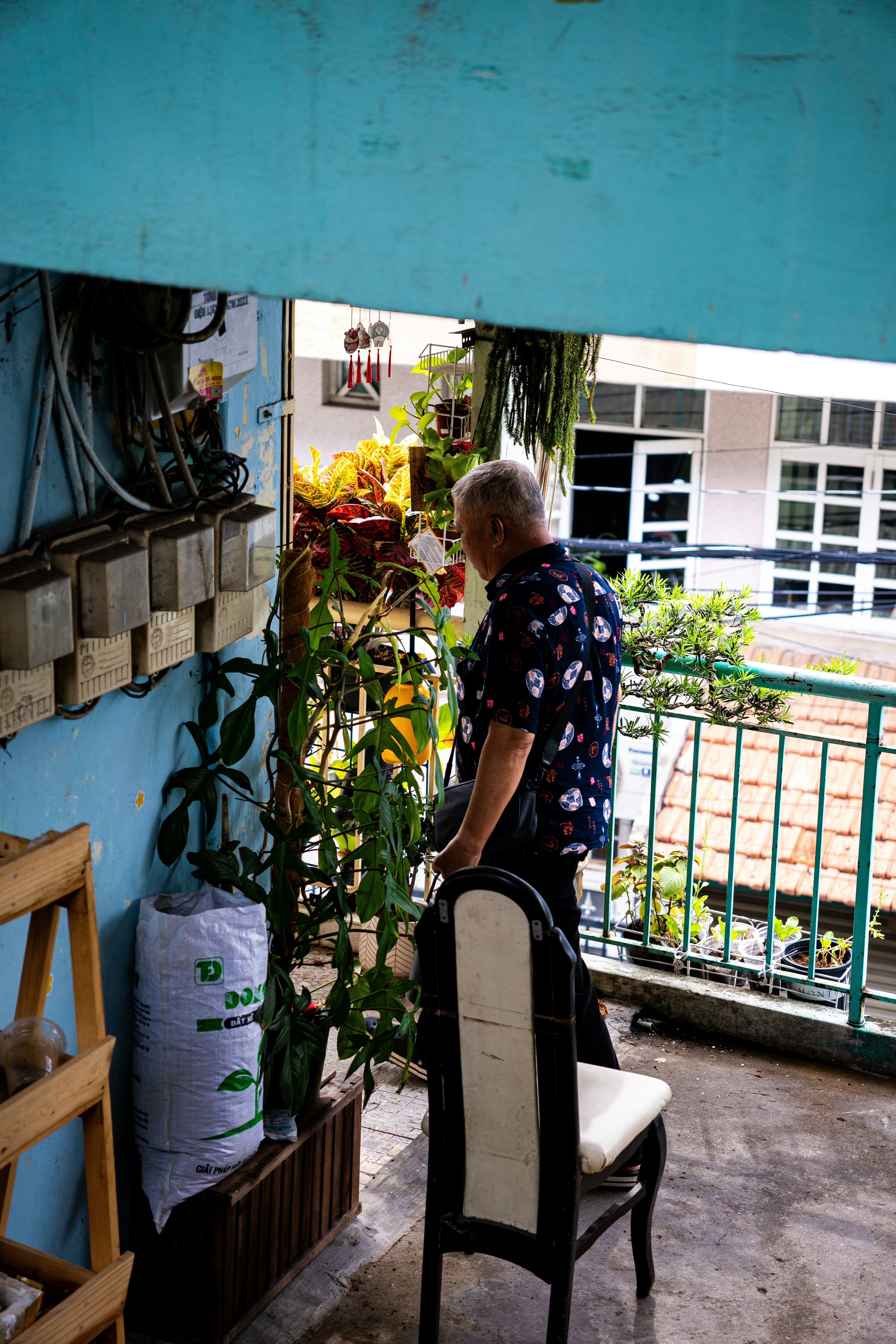 A photograph of a man in a floral shirt tending potted plants on a sunlit balcony, with a turquoise ceiling and a chair nearby. The scene captures urban balcony gardening in a compact outdoor space.