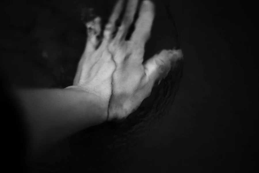 Black-and-white close-up of a hand pressed against a dark, textured surface, with the forearm entering from the left. The low-light scene feels abstract and moody.