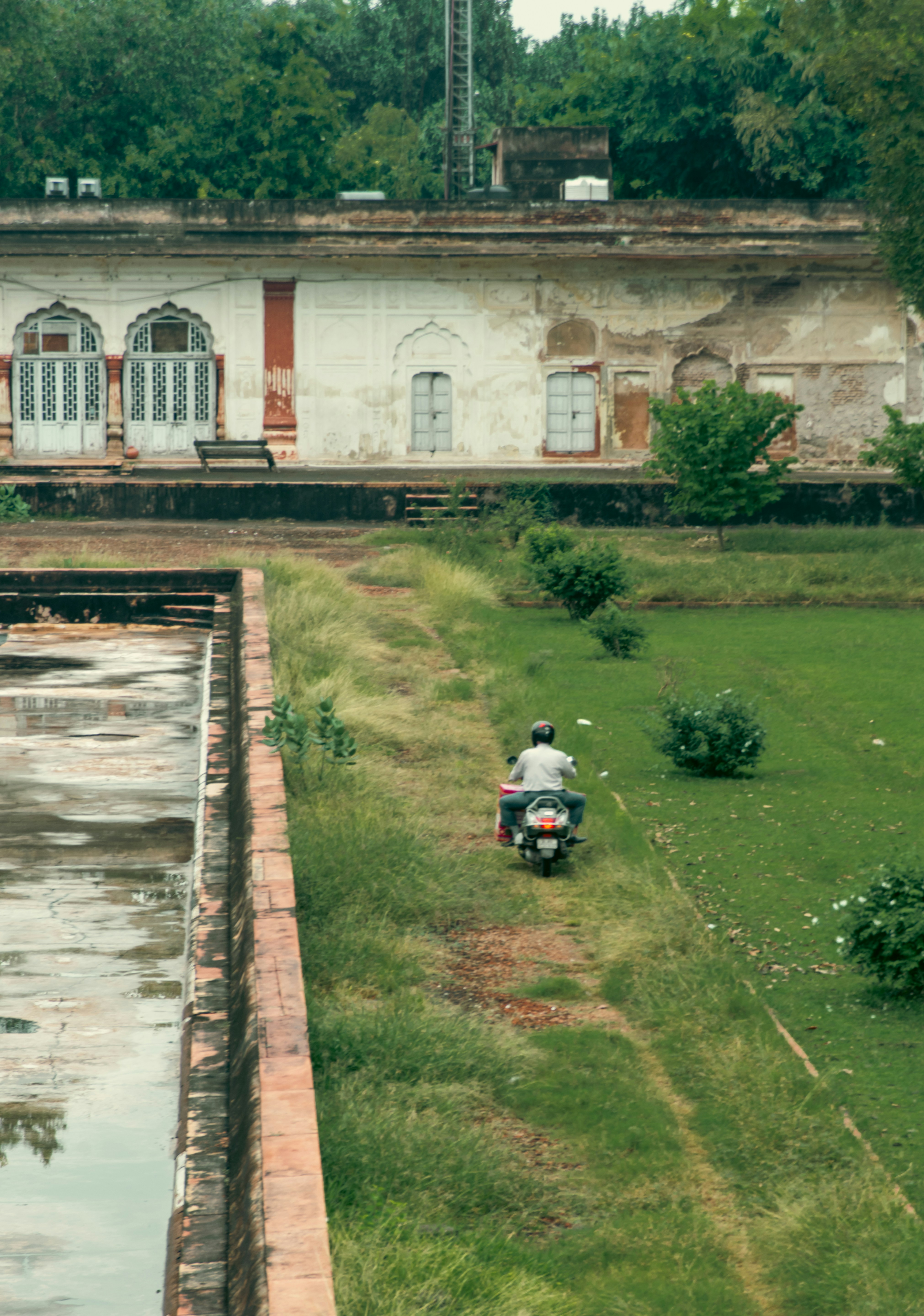 A person rides a scooter along an overgrown path inside the Safdarjung complex in Delhi towards an old weathered building with arched windows and faded plaster, surrounded by greenery after the rain.