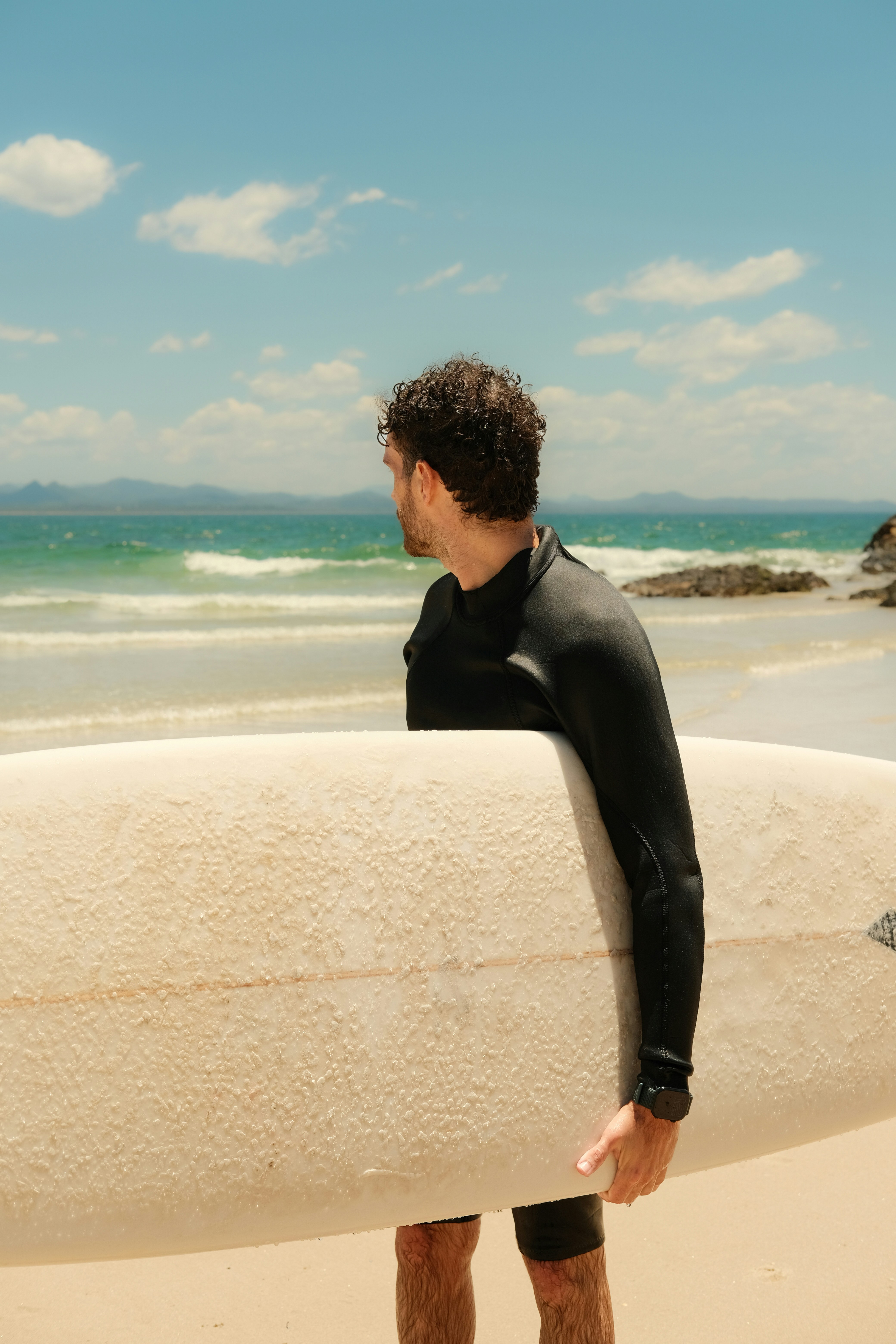 Coastal Pause: A Surfer in Black Wetsuit Steadies a Pale Board Against Sunlit Sand
