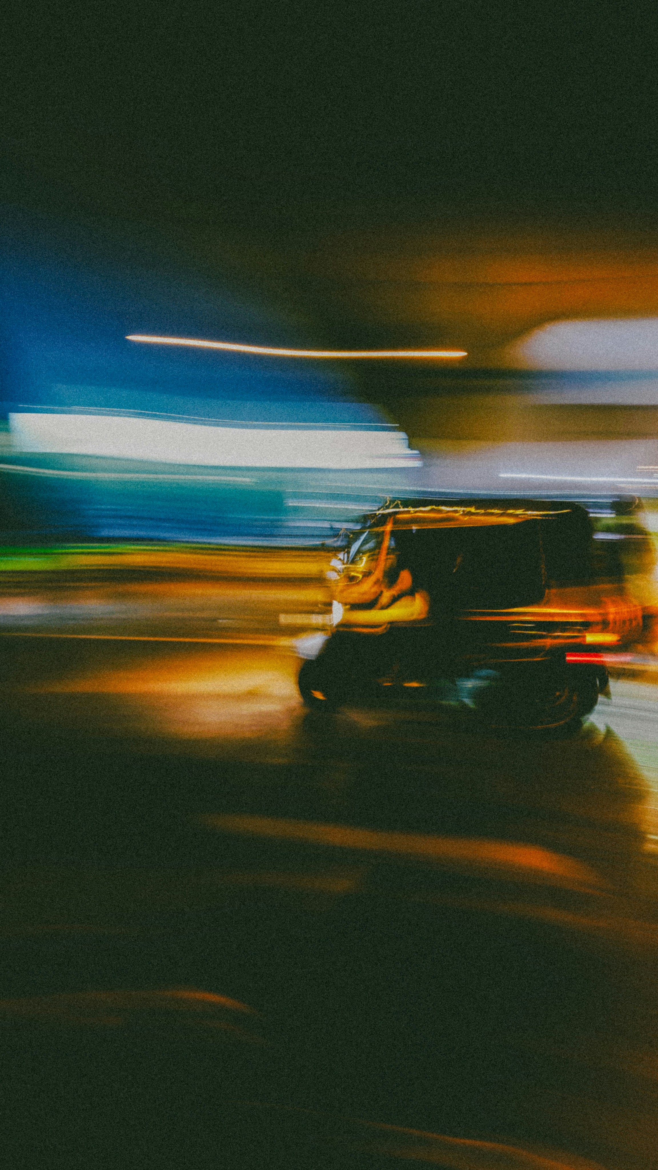 A black van navigates a rainlit cityscape at night, captured with significant motion blur to emphasize speed and urban energy. The scene conveys motion rather than detail, suggesting a dynamic transit moment.