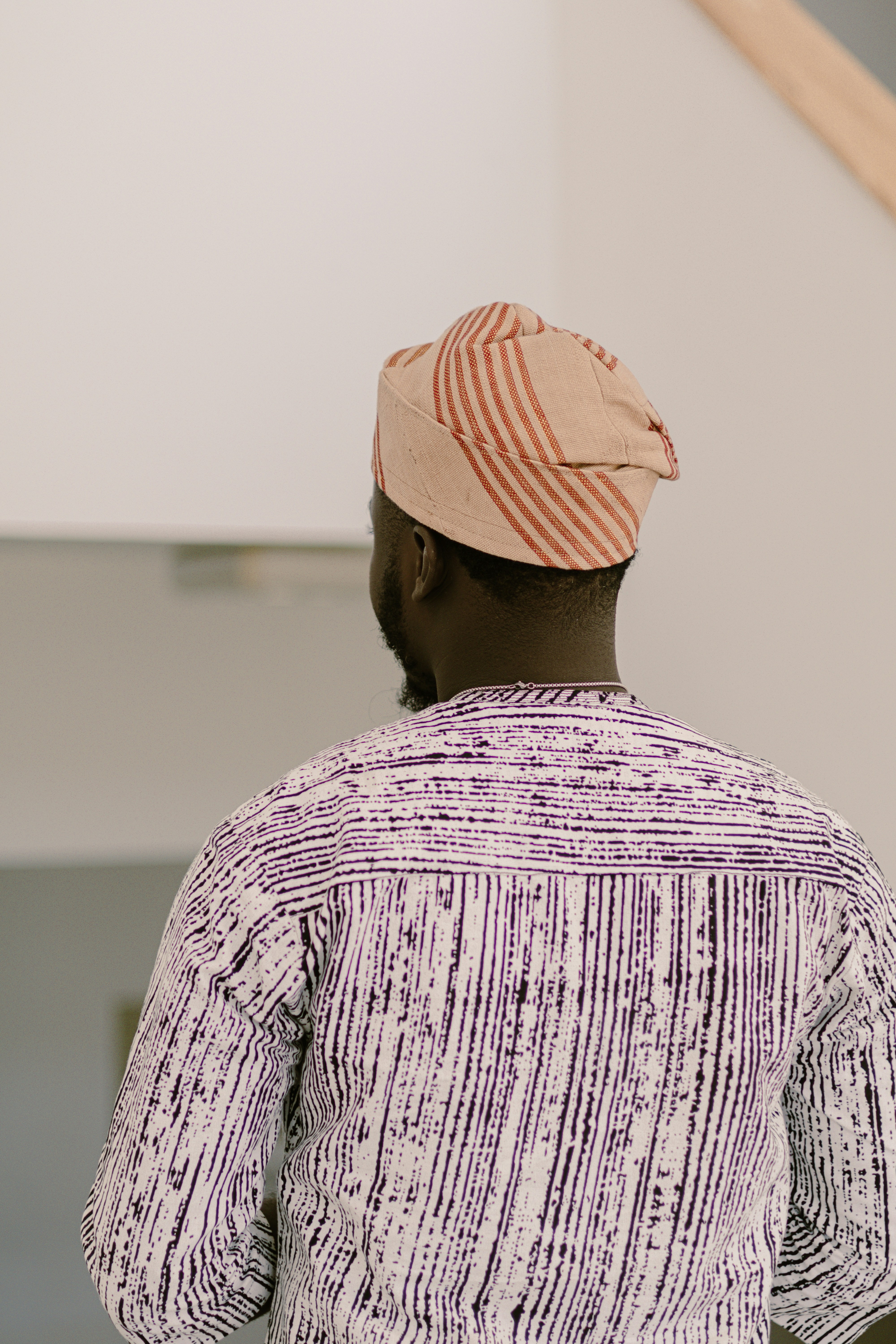 A back view focusing on the striped traditional cap and patterned shirt, showcasing meticulous fabric textures and cultural design. Photographed in warm, diffused light with a simple backdrop for a clean, editorial feel.