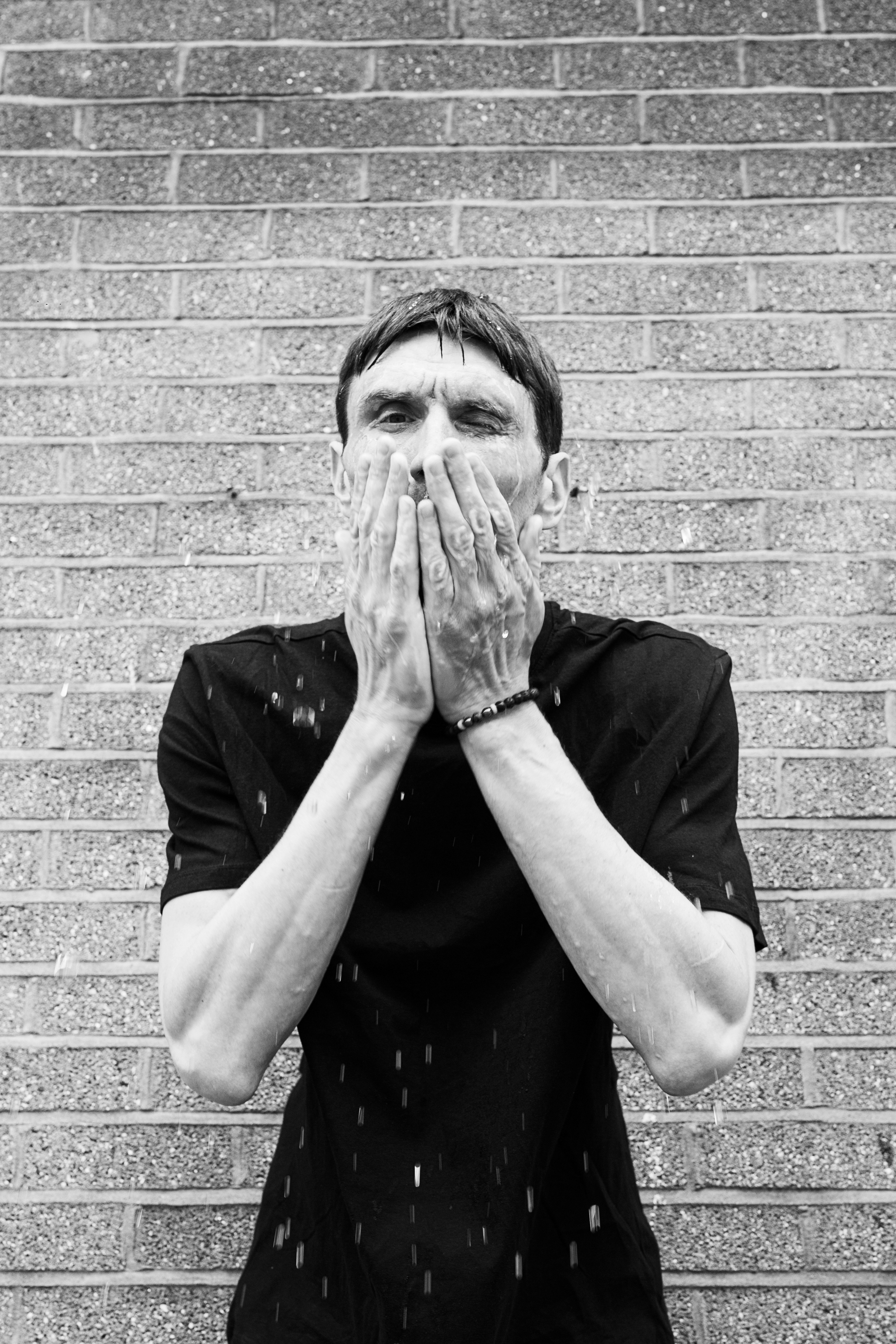 Black-and-white photograph of a man covering his mouth against a brick wall. Droplets speckle the frame, heightening a moment of restrained emotion.