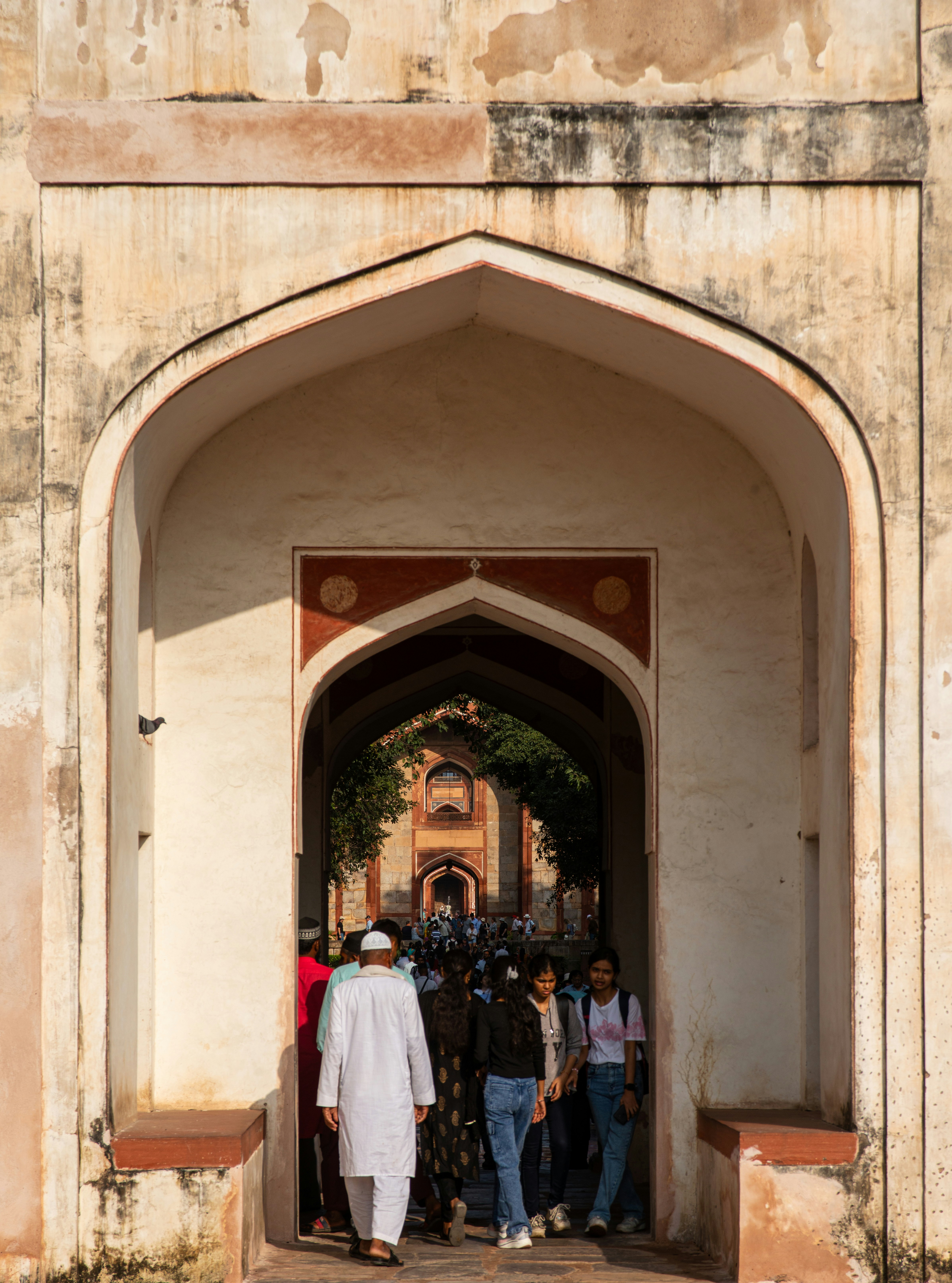 A layered Mughal arch frames a busy walkway at Humayuns Tomb in Delhi. Warm evening light highlights the sandstone details as crowds move toward the monument, creating depth and a sense of movement through the historic gateway.