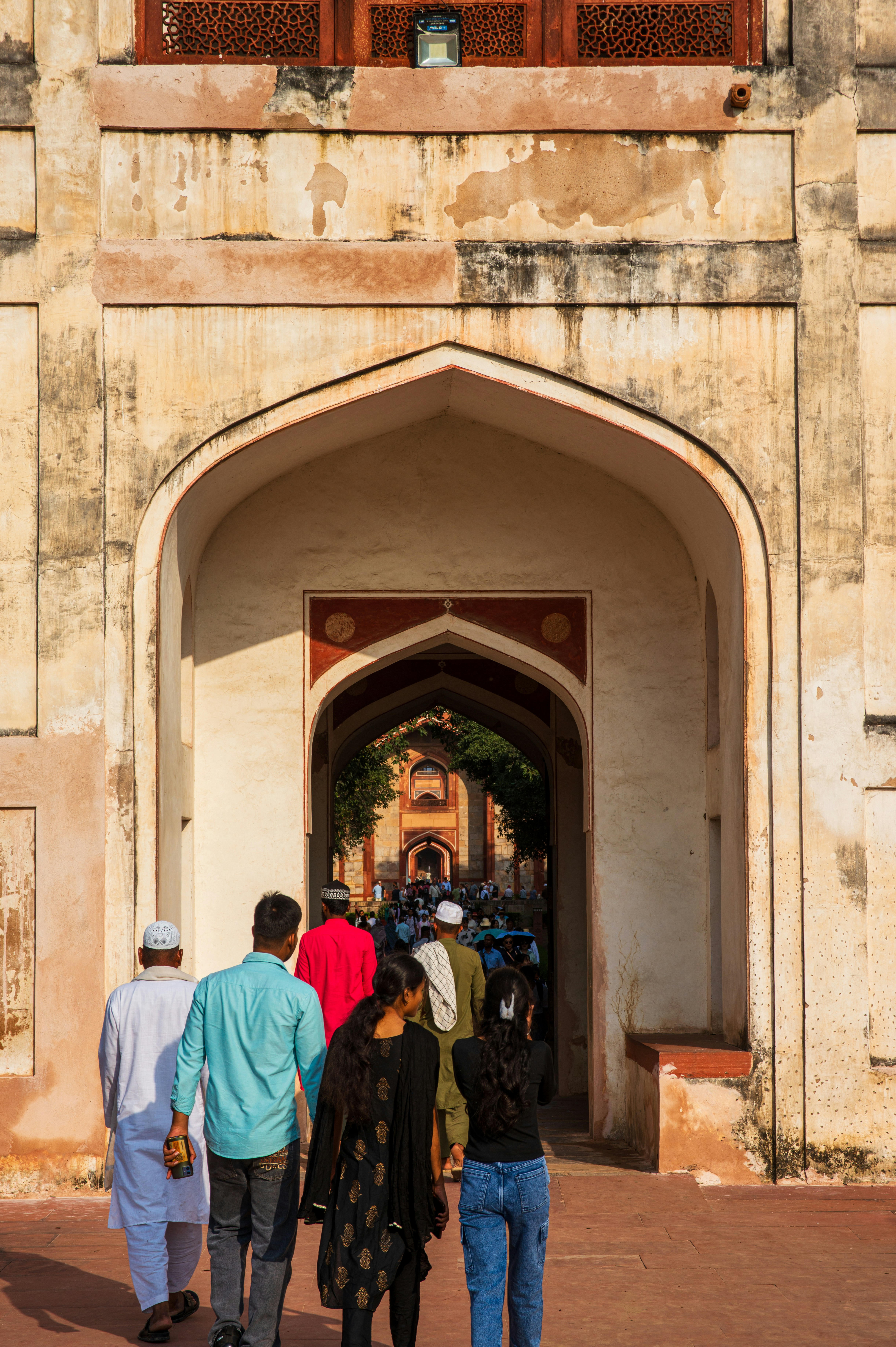 A layered Mughal arch frames a busy walkway at Humayuns Tomb in Delhi. Warm evening light highlights the sandstone details as crowds move toward the monument, creating depth and a sense of movement through the historic gateway.