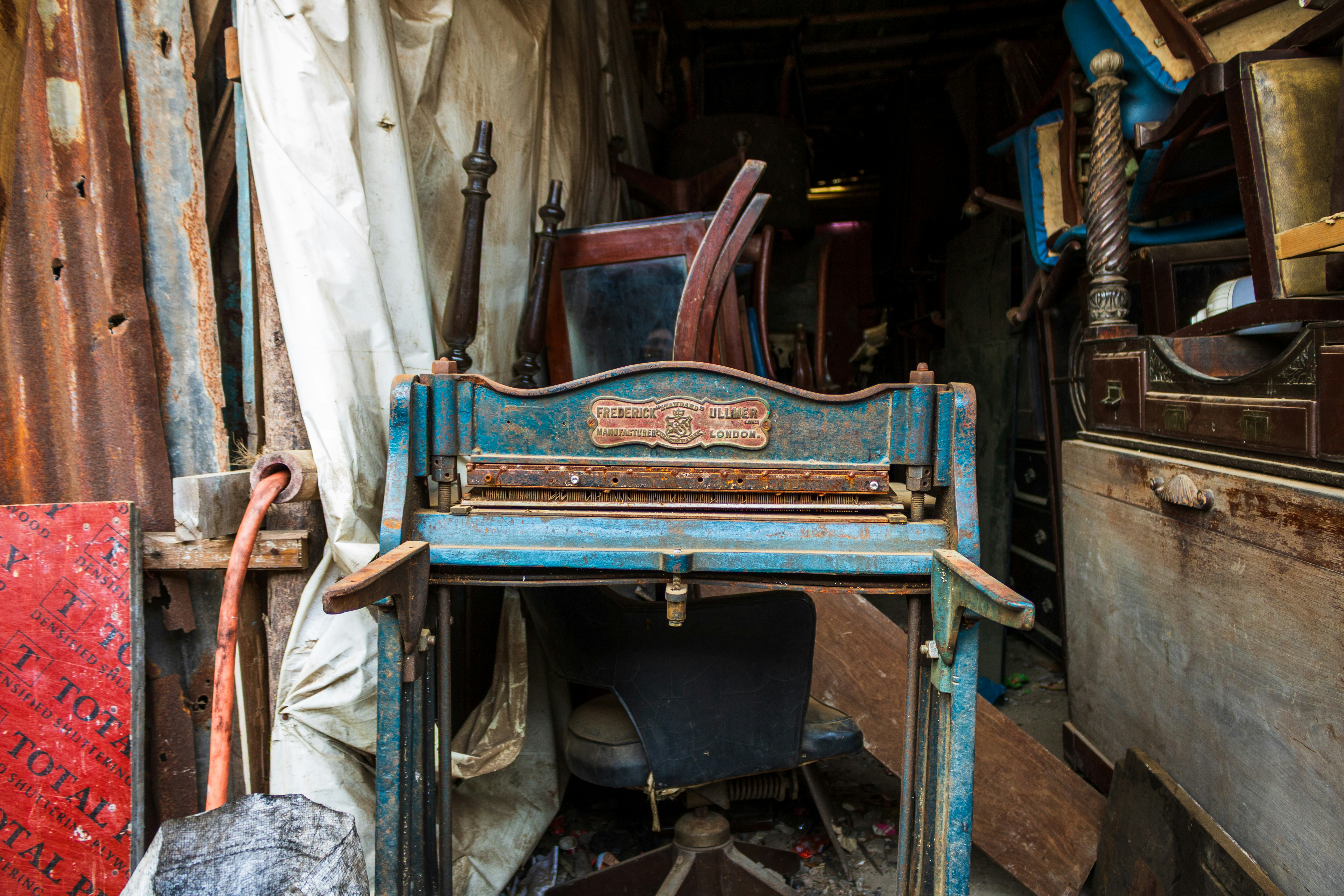 A blue, vintage "Frederick Ullmer, London" machine used for pressing or cutting. The rough, peeling paint and rust on the machine contrast with the surrounding textures: corrugated, rusty metal sheeting, draped white canvas, and stacked wooden furniture. This rich composition is excellent for editorial features on antiques, craftsmanship, or urban salvage, or as a dense background to convey a sense of history, complexity, and organized chaos in a design project.