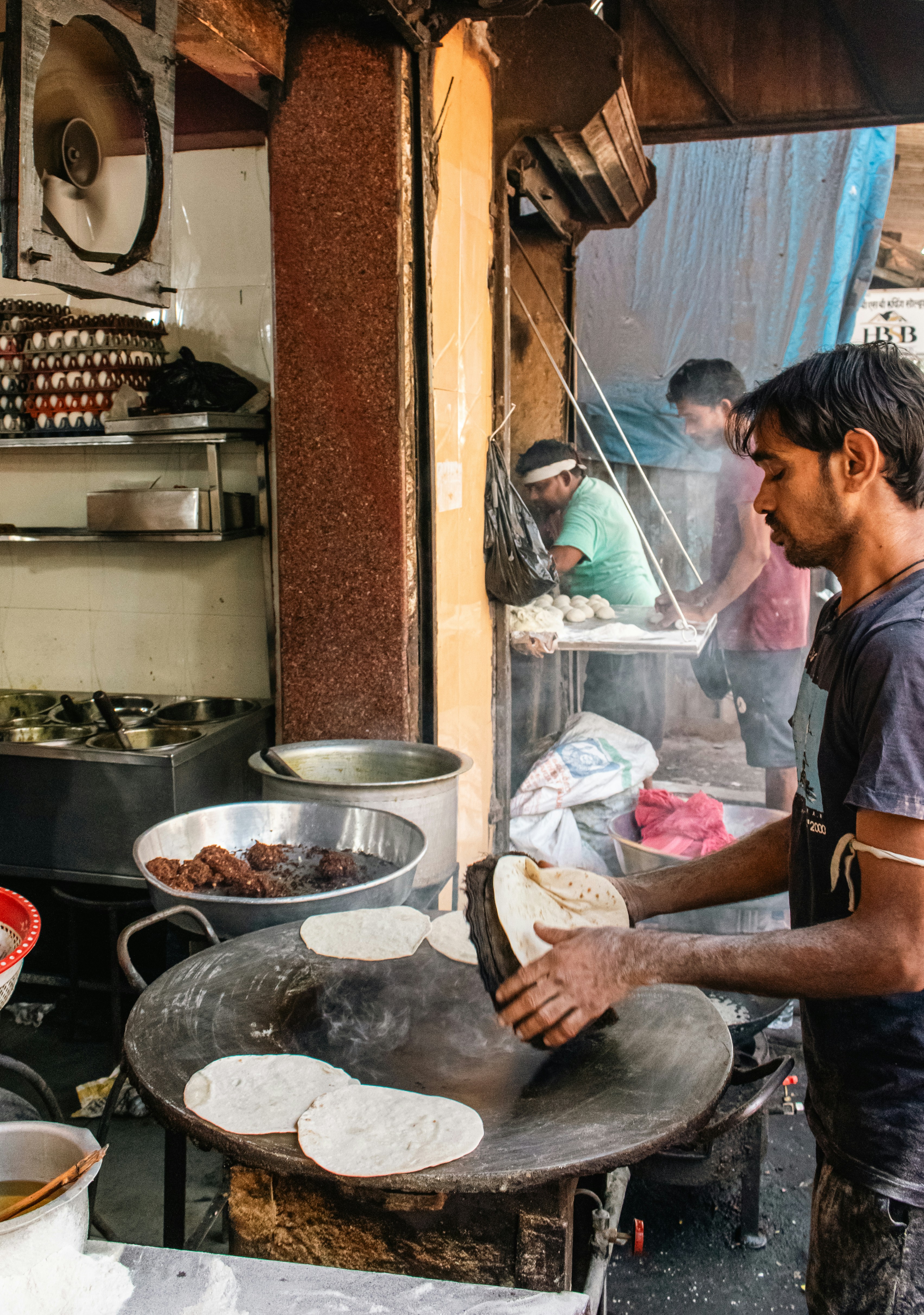 A street vendor in Mumbai preparing Indian flatbread on a large, blackened tawa or griddle. The vendor's arms, dusted with flour, handle a partially cooked, round bread, emphasizing the texture of the dough against the rough iron surface. The ambient light highlights the simple, rustic process of cooking, making it ideal for content related to street food, culinary traditions, travel in India, or authentic cooking blogs and articles, conveying a sense of immediacy and cultural authenticity.