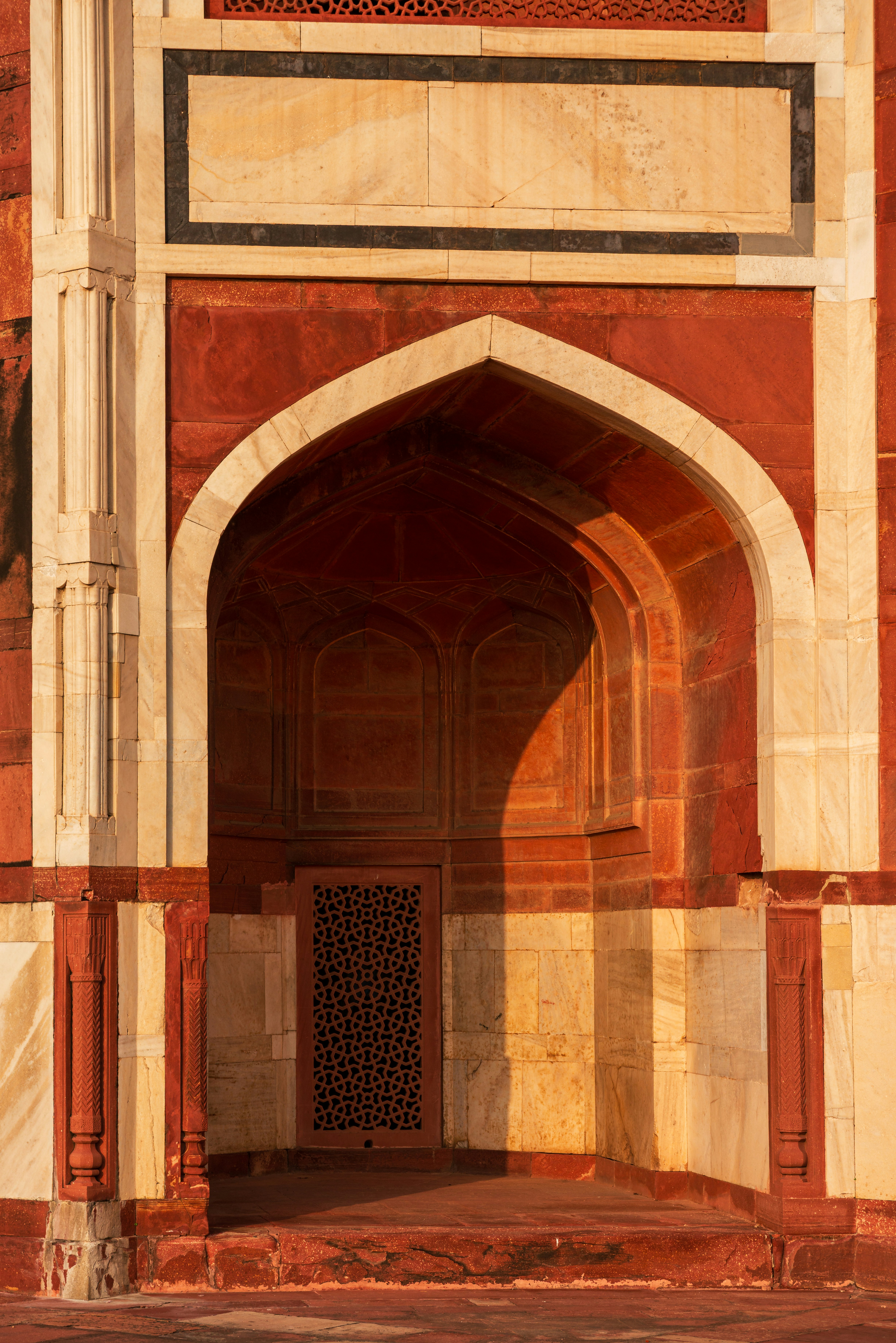 A vertical close-up captures a recessed arched niche (iwan) on the facade of a Mughal monument, likely Humayun's Tomb, illuminated by warm, low-angle light. The structure is primarily red sandstone with contrasting vertical white marble panels and dark stone trim. The arch frames a small, dark recess containing a stone lattice screen or jali at ground level.