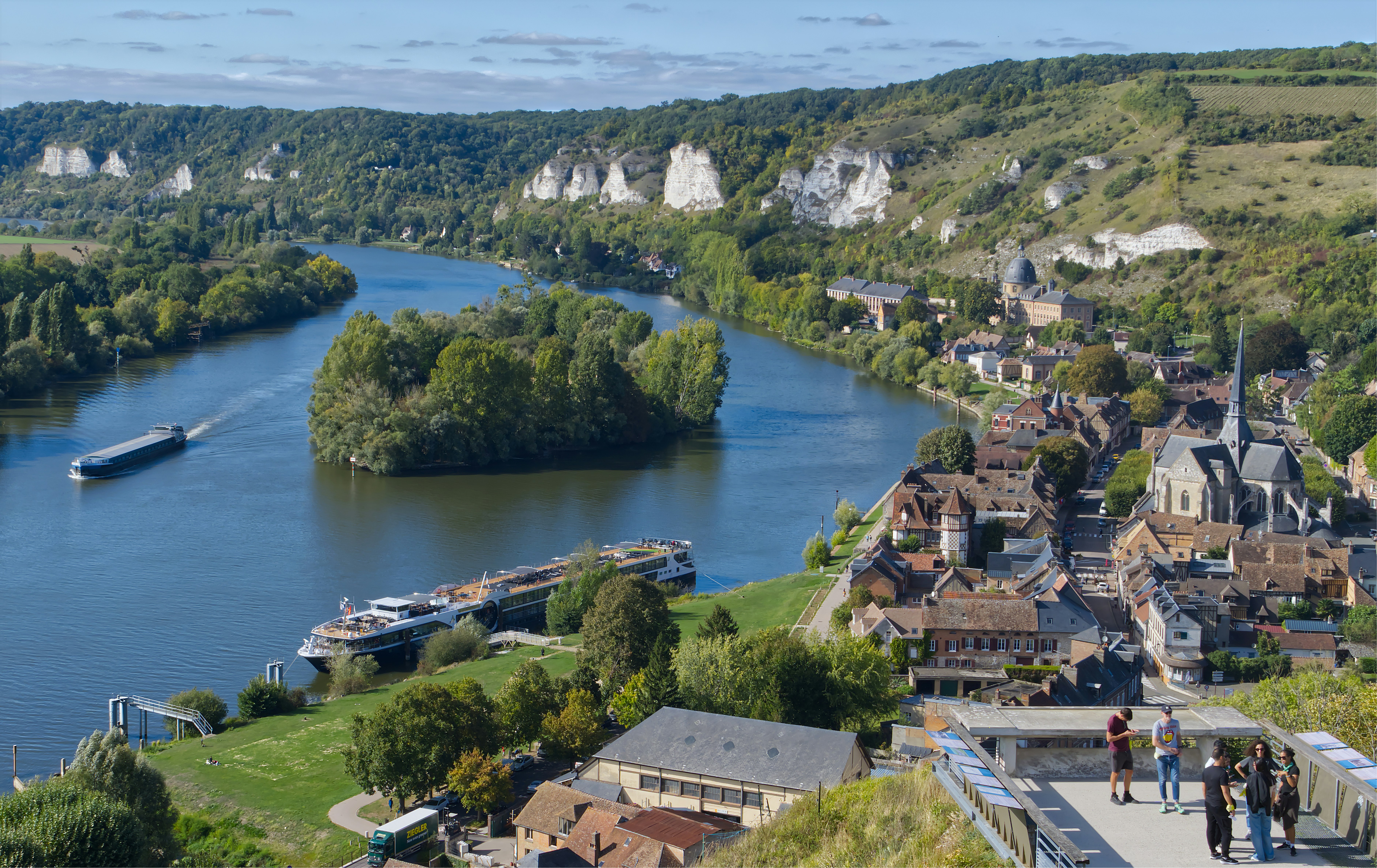 Aux portes de la Normandie, la boucle de la Seine aux Andelys déploie l’un de ses plus beaux décors naturels, où le fleuve, large et paisible, épouse la forme des collines avant de se heurter aux impressionnantes falaises de craie. Ces géants blancs, véritables signatures géologiques de la région, dominent le paysage de leur silhouette majestueuse. Au pied de ces parois, le village des Andelys s’étire le long des berges, avec ses maisons traditionnelles, ses ruelles étroites et son église qui se fondent harmonieusement dans ce cadre naturel préservé. Les bateaux de croisière, glissant sur le fleuve, permettent d’apprécier la hauteur vertigineuse des falaises et la douceur des coteaux boisés qui les entourent.