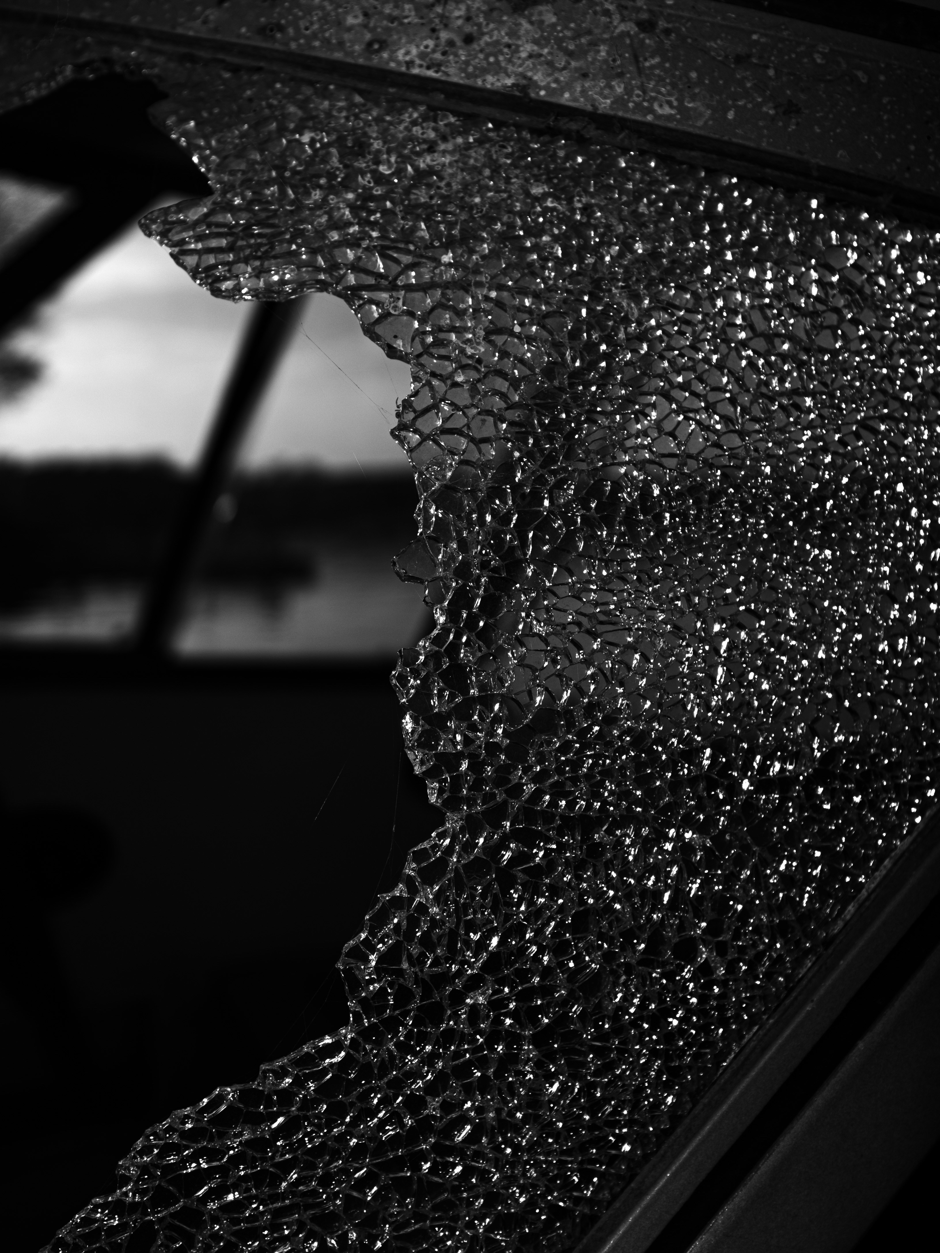 Monochrome close-up of a cracked car window, with jagged glass patterns catching light against a dark interior and blurred background.
