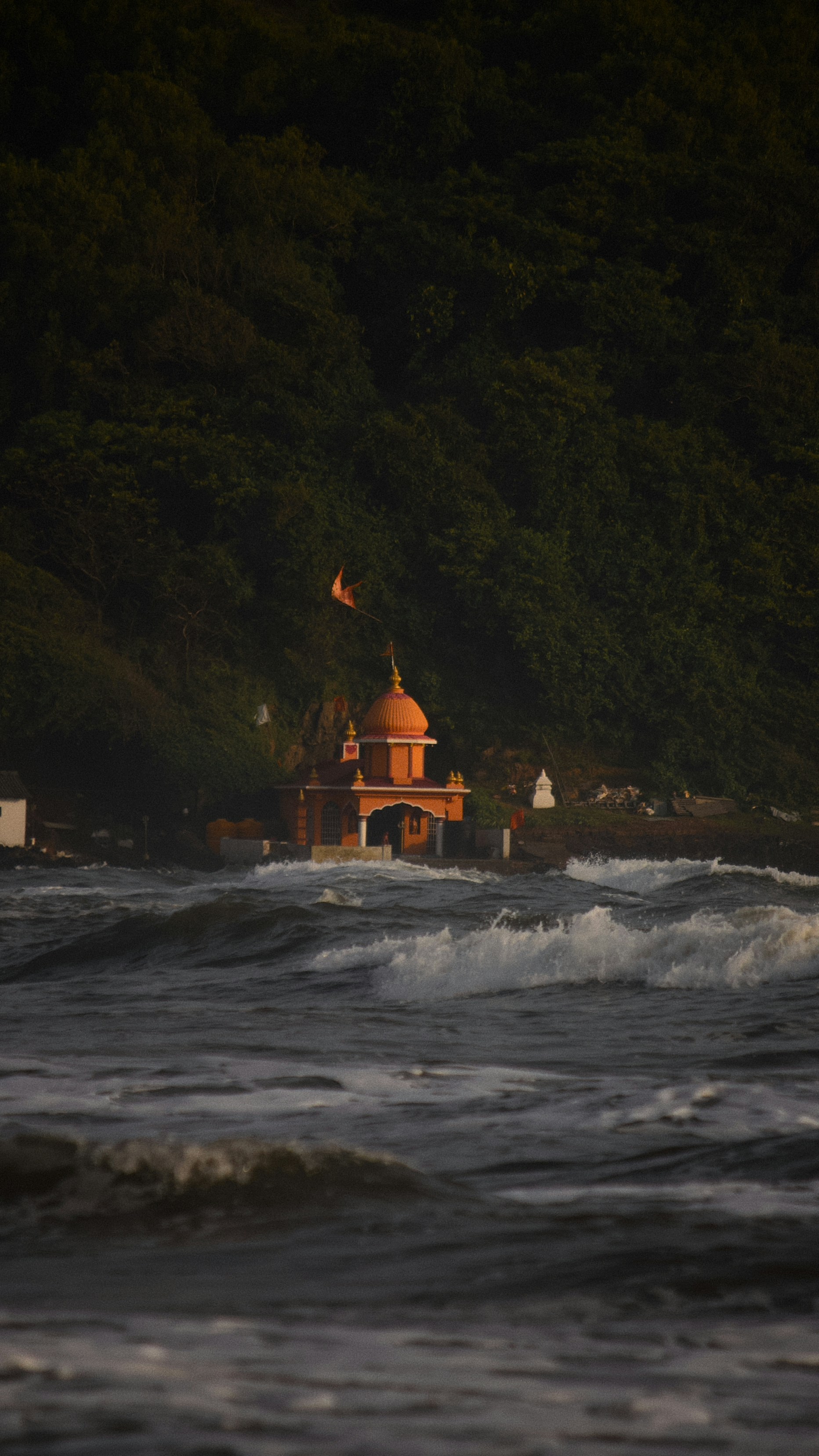 A small coastal temple in Goa, India glows with warm orange tones against a backdrop of dense tropical greenery. Soft evening light highlights the dome while powerful ocean waves crash in the foreground, creating a serene yet dramatic atmosphere typical of Goa’s monsoon coastline.
