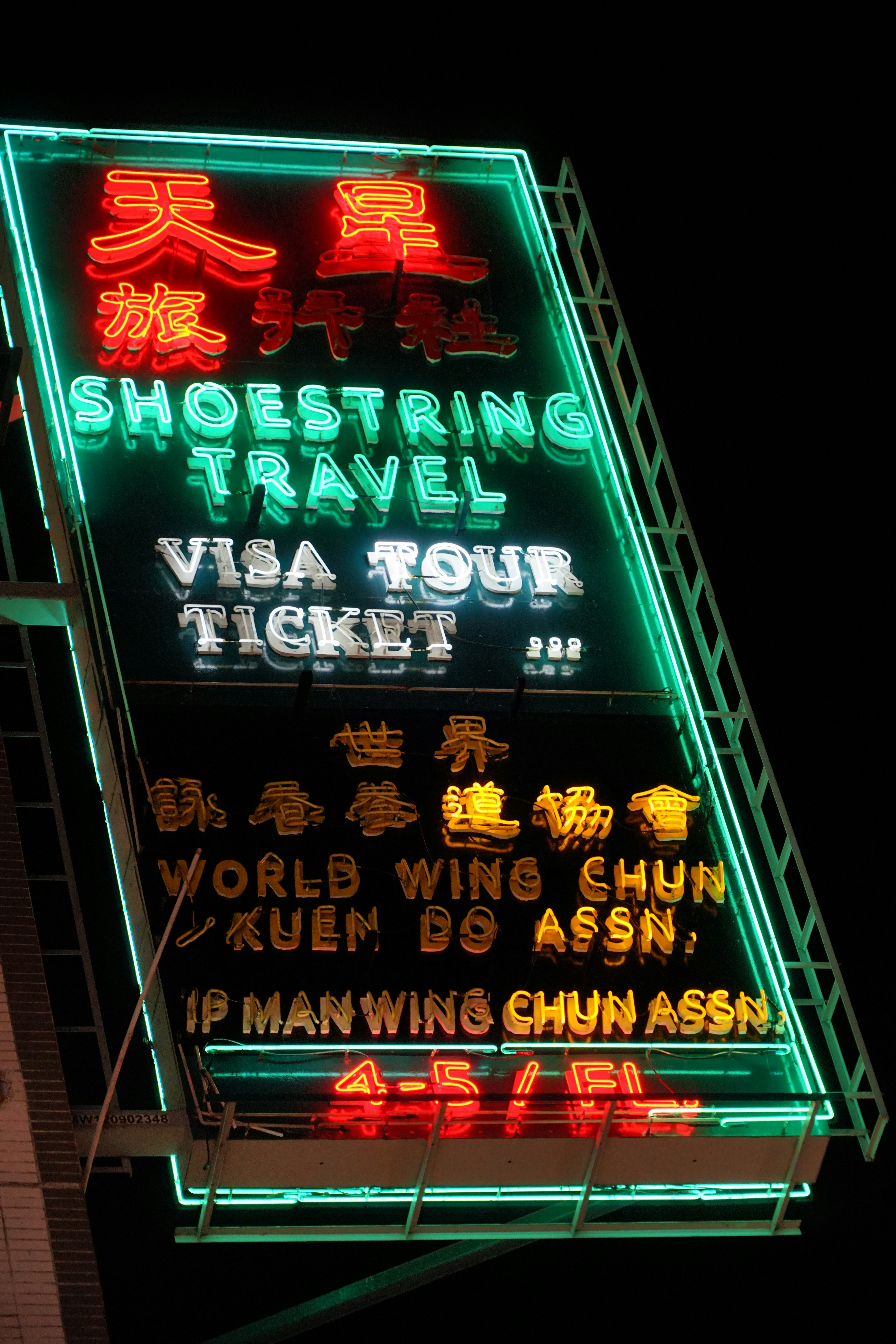A realistic neon sign installation adorns the side of an office building in Hong Kong at night, featuring two distinct neon signboards with clear, spelled-out text: one reads "world wing chun m global" and the other displays "i p man winston chun as travel ticket". The neon lights emit vibrant hues of green, red, yellow, and orange, illuminating the building’s exterior in the nighttime darkness. Below the main neon signs, additional text such as "4-5-f/" is visible, and the entire neon sign setup is rendered with a highly realistic appearance, capturing the vivid glow and detailed lettering ch