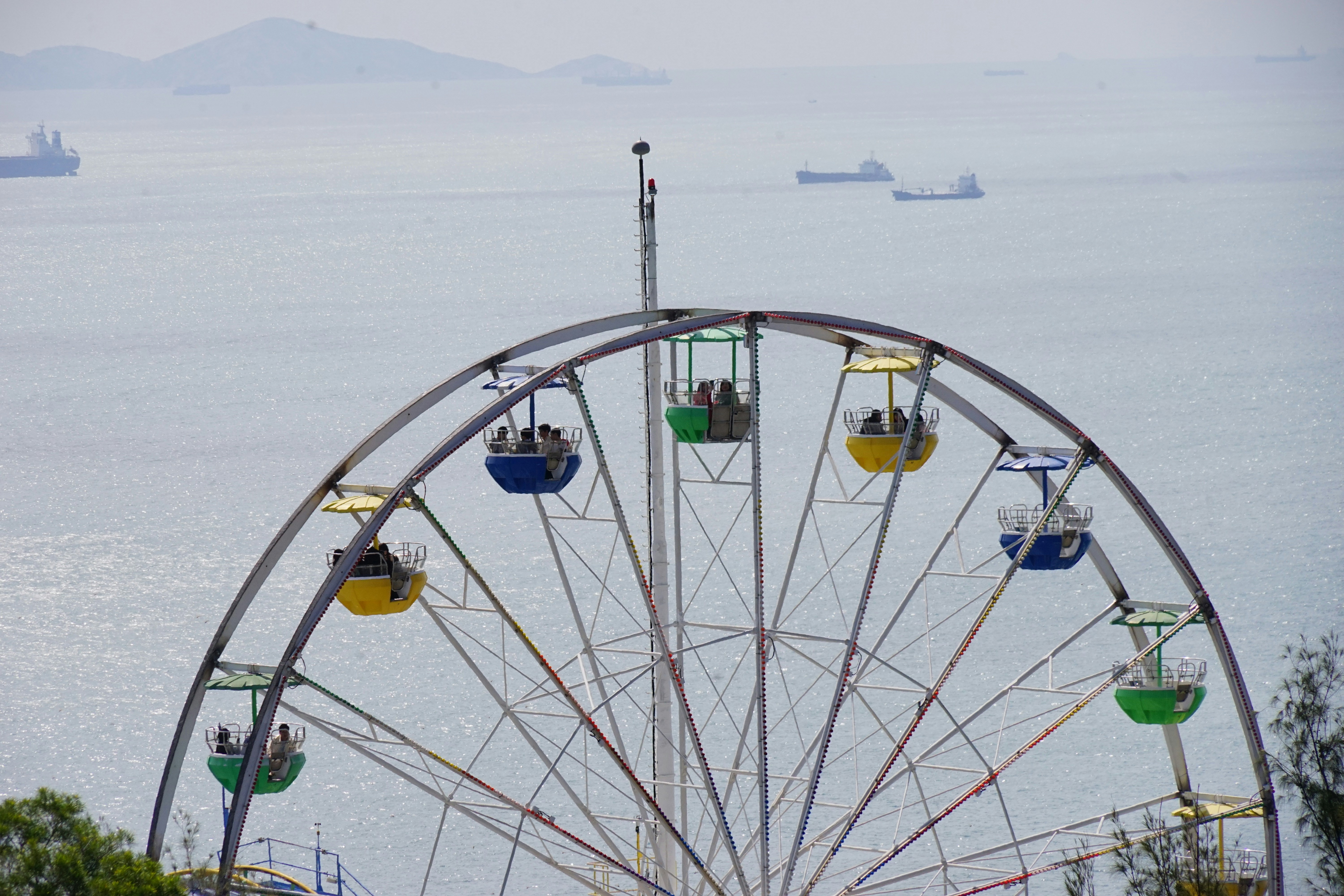 A vibrant, multicolored ferris wheel featuring green, blue, and yellow carriages rises prominently against the backdrop of the sea at Pohang's seaside. Inside one of the ferris wheel cabins, three individuals are seated, smiling as the cabin ascends to roughly the halfway point of the wheel’s rotation. In the distance, along the horizon, several cargo ships are visible on the water. The photograph is captured from a top-angle perspective on a bright, sunny day, highlighting the cheerful coastal scene and the vivid hues of the ferris wheel against the calm sea.