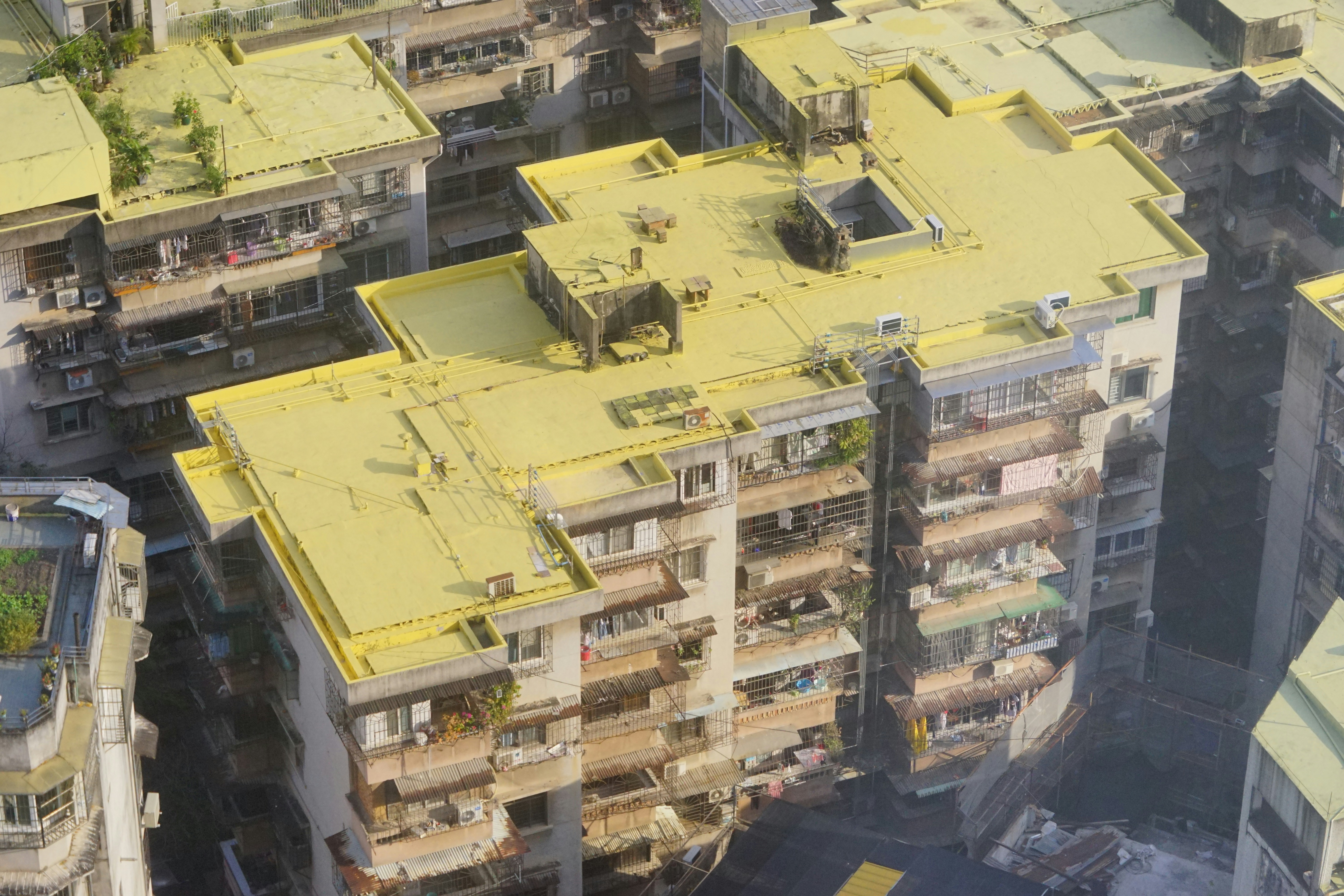 This is an aerial view of dense multi-story residential buildings (typical of urban neighborhoods in many East Asian cities) characterized by distinct yellow-painted rooftops—a notable feature that may serve as a protective coating (e.g., waterproofing) or aesthetic uniform. Key details include: Building features: Aged concrete facades, balconies (some with potted plants and hanging laundry, indicating residential use), and small rooftop structures (vents, pipes). Environment: Clustered apartment blocks, creating a high-density urban residential landscape; subtle greenery (small plants) is vis