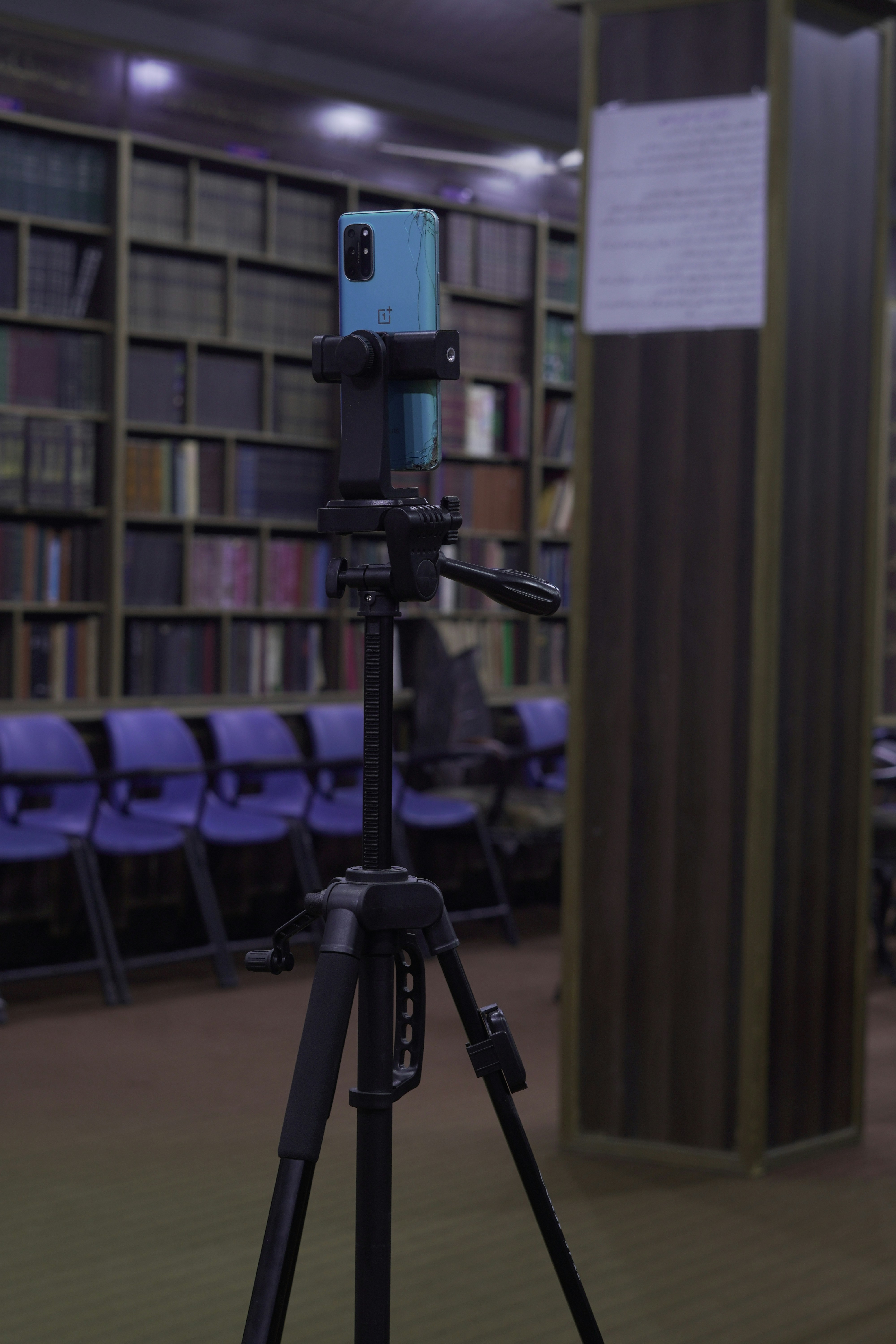 Phone on Tripod in a Quiet Library A smartphone mounted on a tripod stands in the middle of a quiet library hall, surrounded by shelves filled with books. Soft indoor lighting and empty chairs create a peaceful academic atmosphere, capturing a moment of stillness before recording begins.