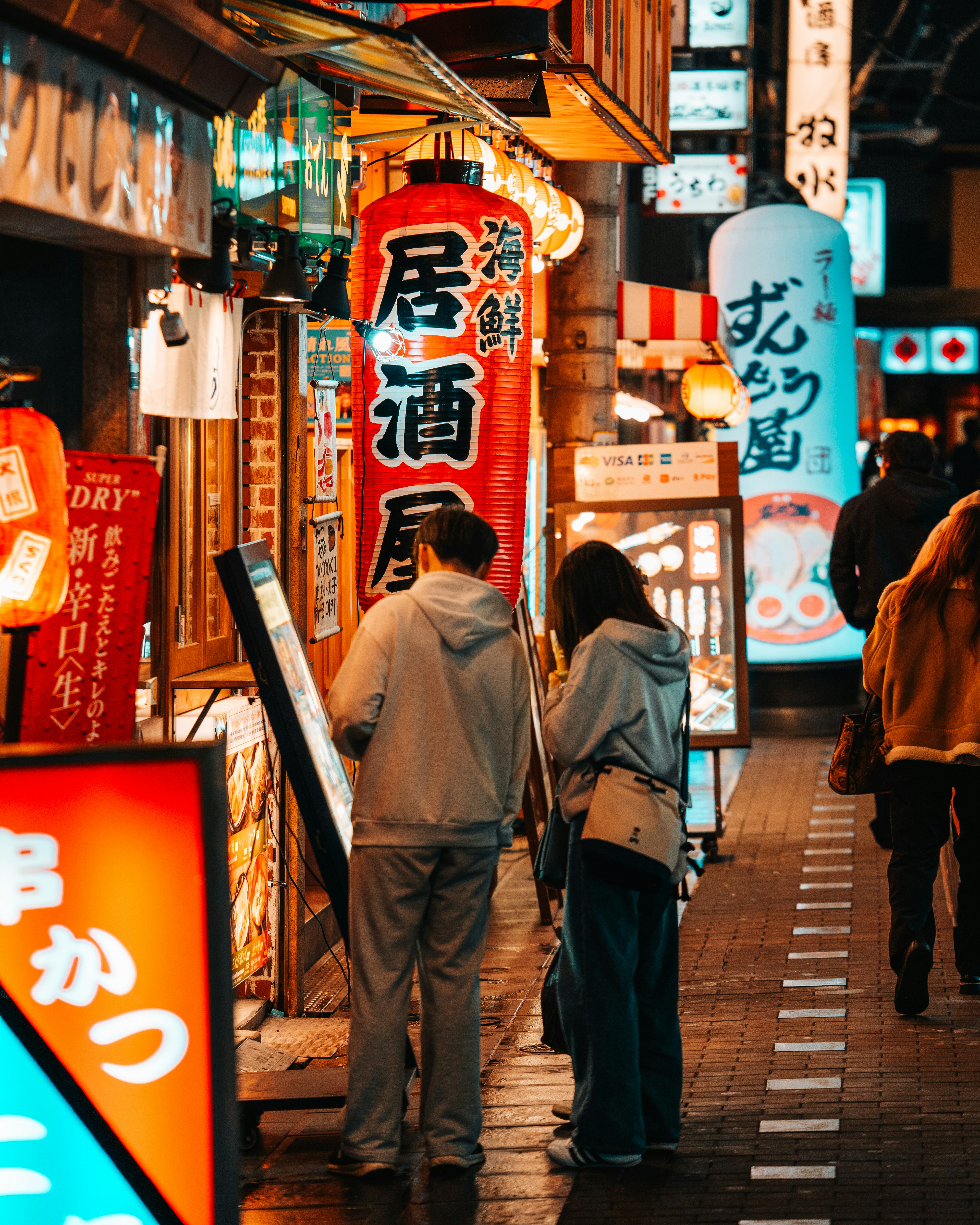 A young couple stands at the entrance of a restaurant in Dotonbori, carefully studying the menu under the glow of neon signs. The shot captures an intimate moment of decision-making and the atmosphere of the bustling night district where culinary life thrives.
