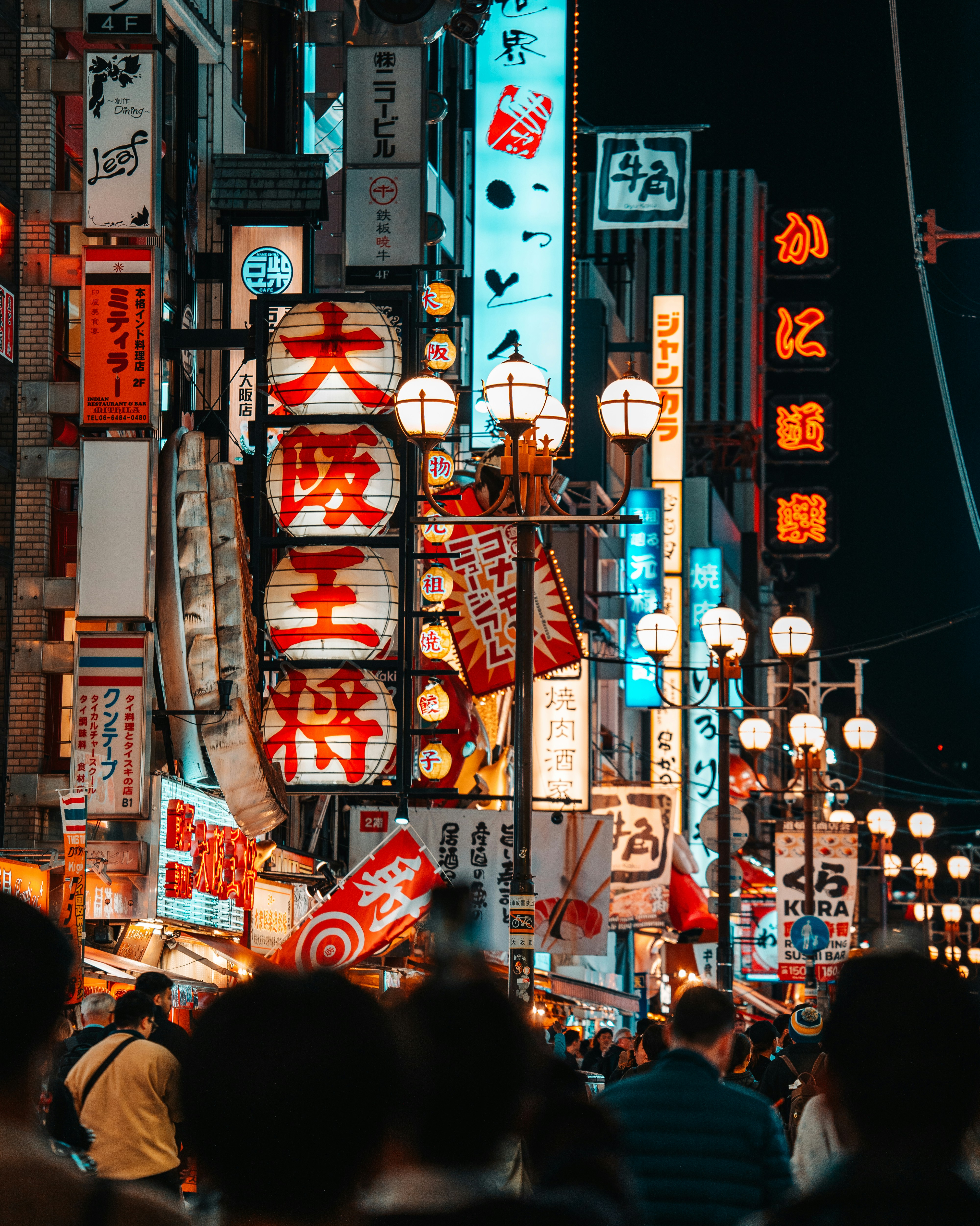 An atmospheric night shot in Dotonbori, where a traditional paper lantern and vibrant neon signs create a colorful composition. In the foreground, an artistically blurred crowd conveys the dynamics and energy of Osaka's famous nightlife district.